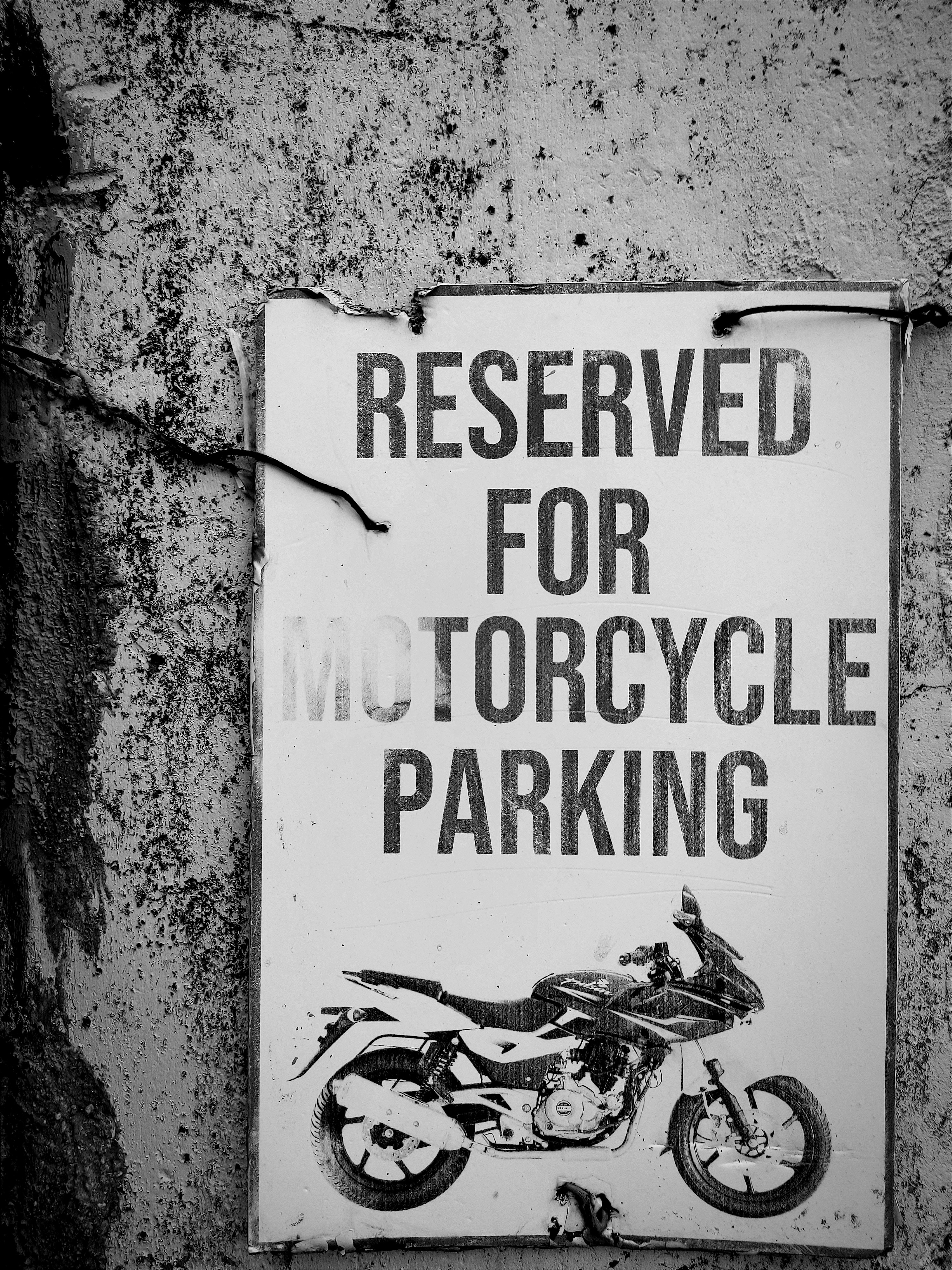 Black-and-white photograph of a weathered sign reading 'Reserved for Motorcycle Parking' mounted on a textured wall, with a motorcycle illustration at the bottom.