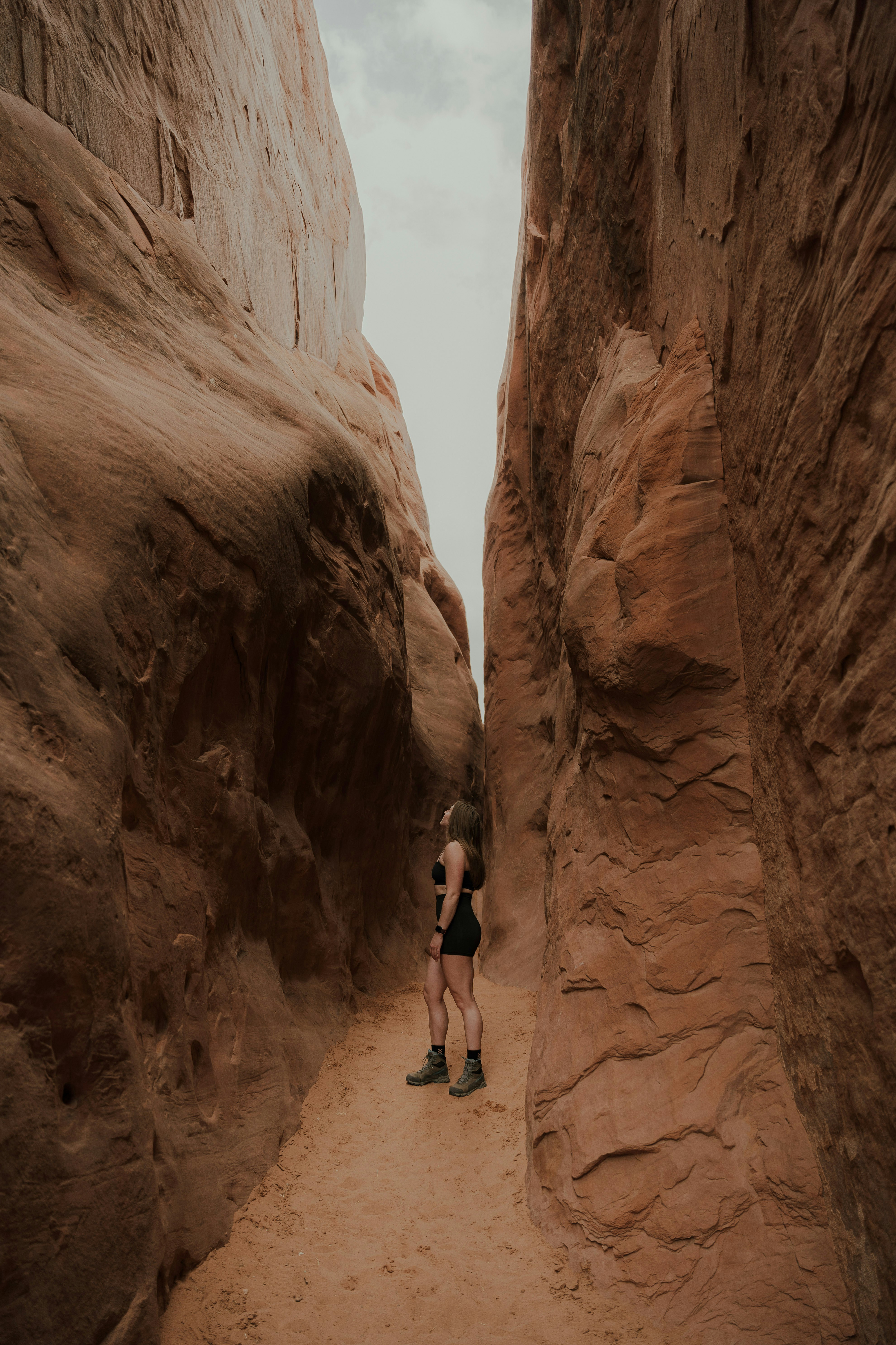 Hiker gazing upward in a narrow canyon, surrounded by towering sandstone walls. The sandy floor leads deeper into the natural formation.