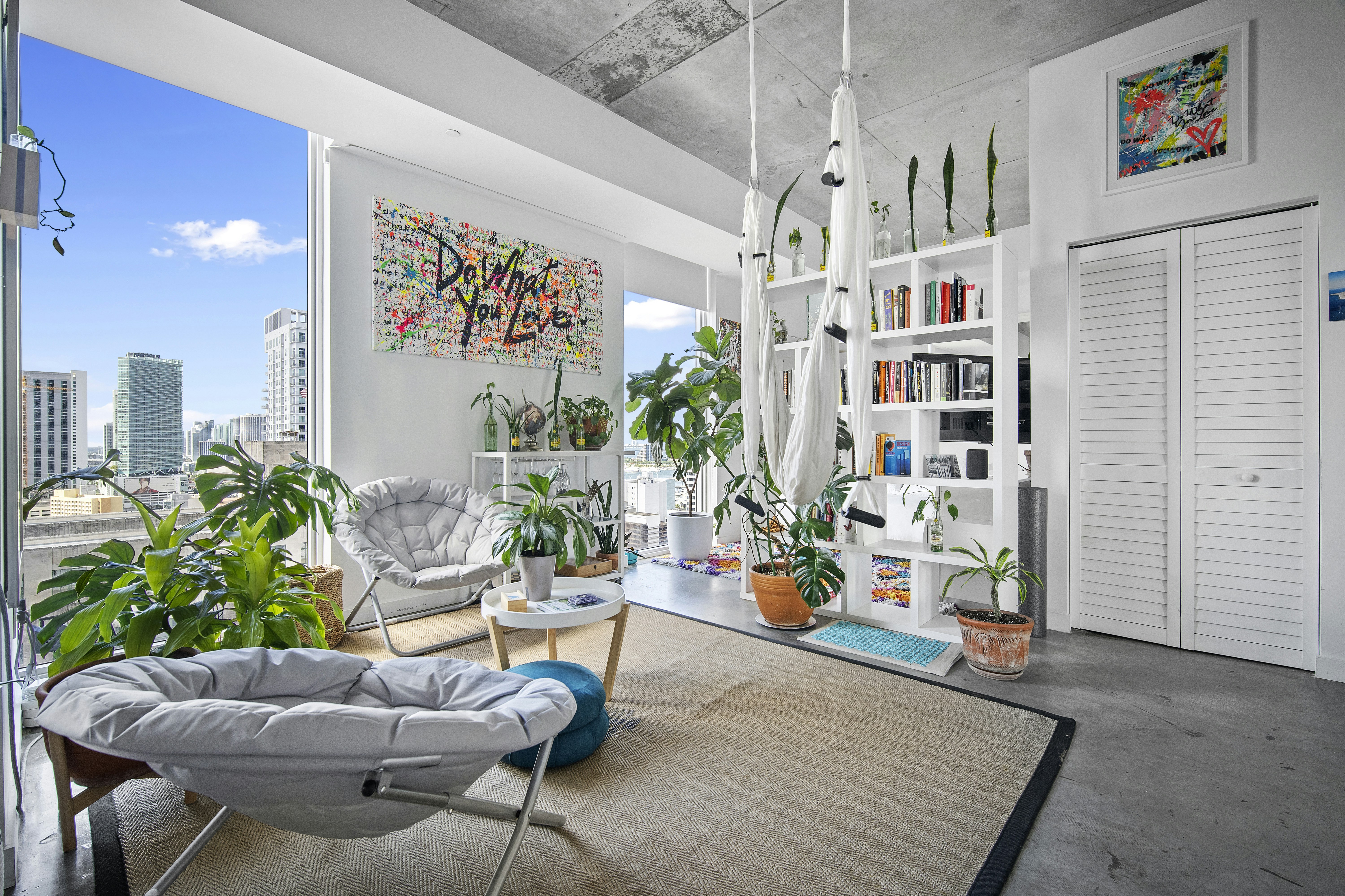 Modern living room with lush plants, hammock chairs, and a vibrant cityscape view through large windows.