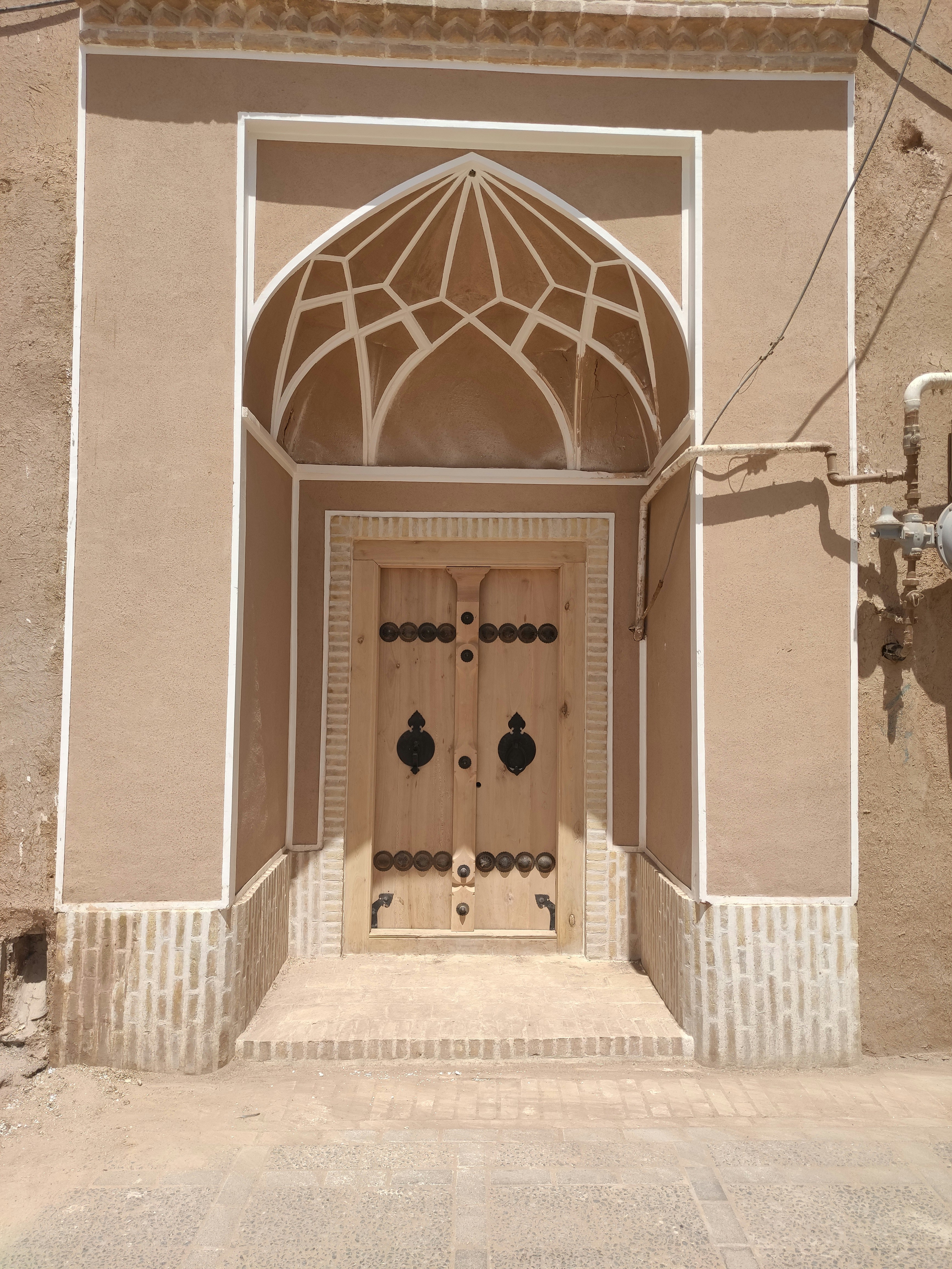 Photograph of a symmetrical doorway with an ornate arch and wooden door set in sunlit beige plaster walls.