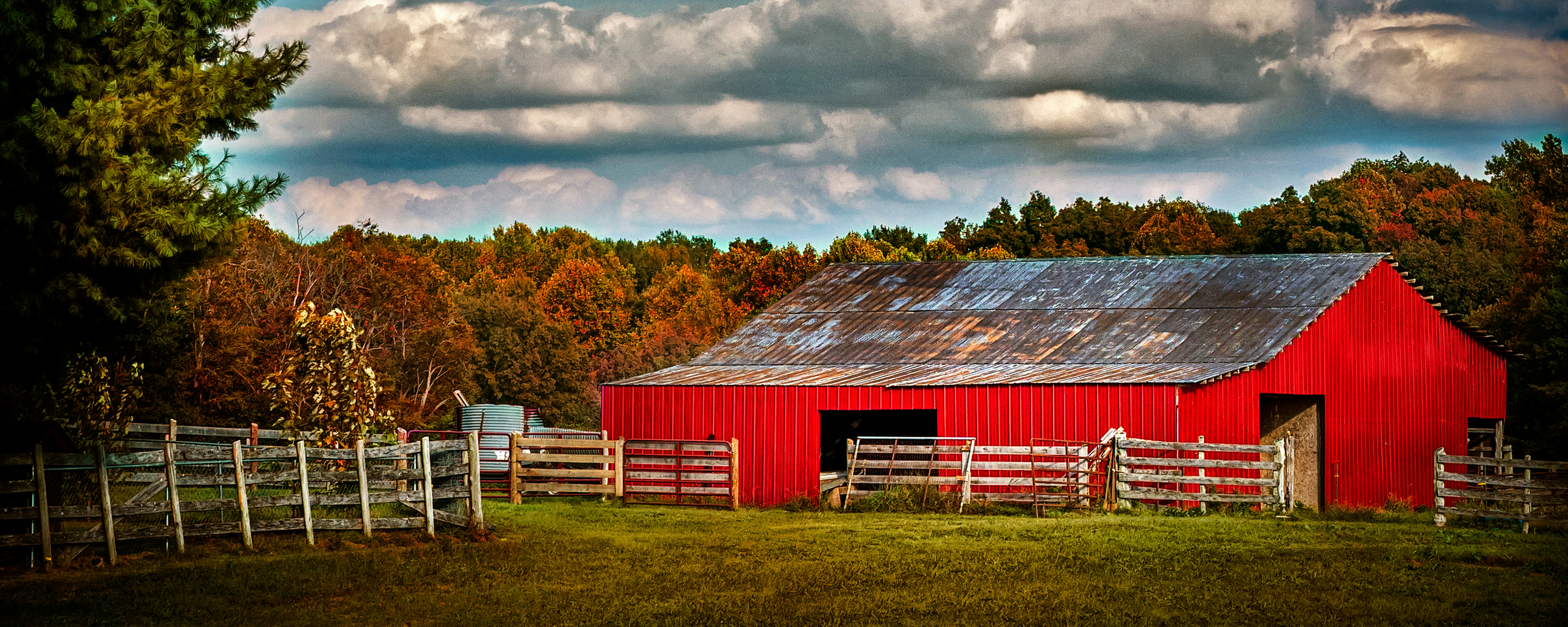 Autumn landscape photograph featuring a red barn with a weathered metal roof, flanked by white fences. The backdrop reveals a line of trees in warm fall colors.