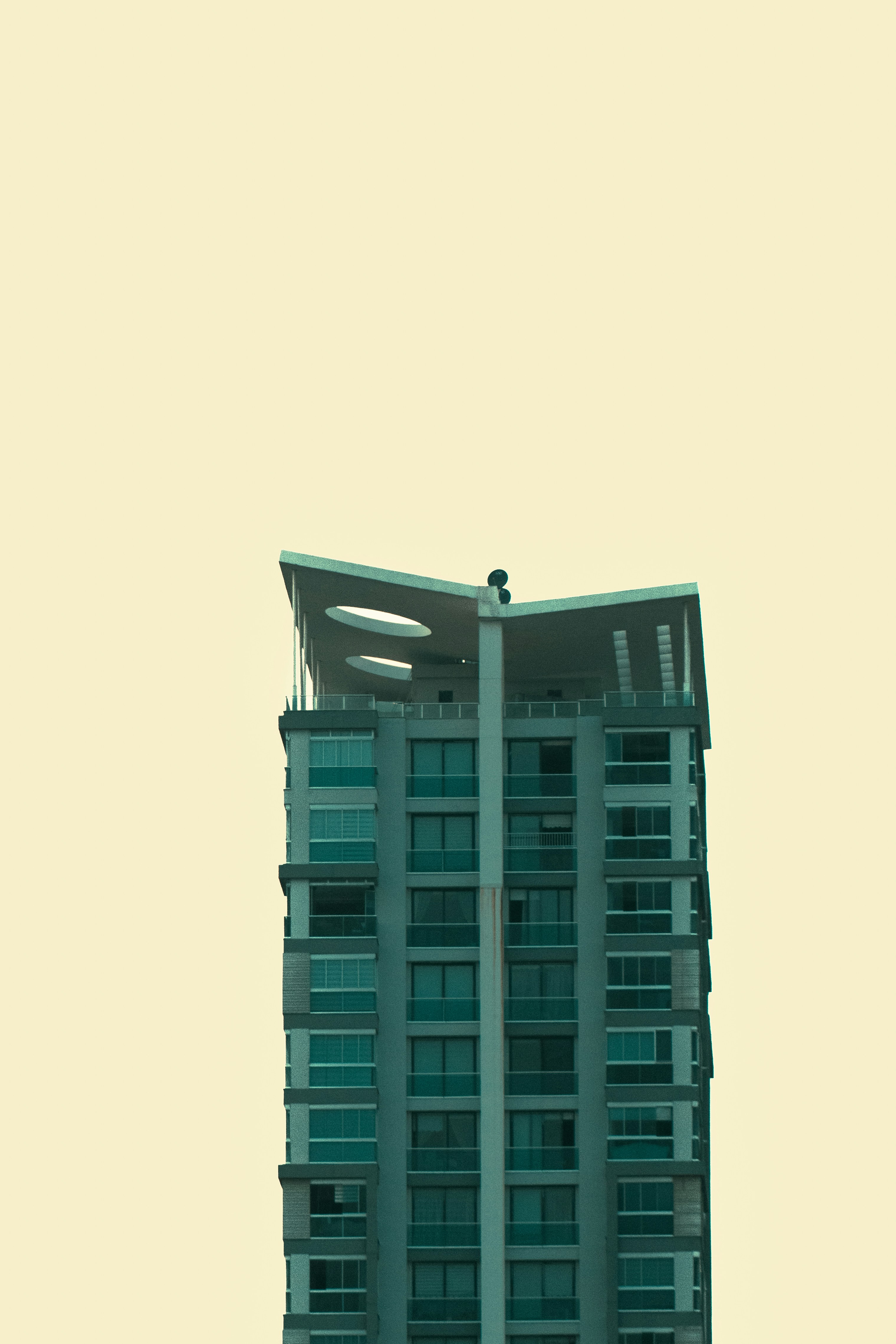 Teal-green high-rise tower against a pale cream sky, isolating the architectural form. Its vertical rhythm of balconies emphasizes geometry.