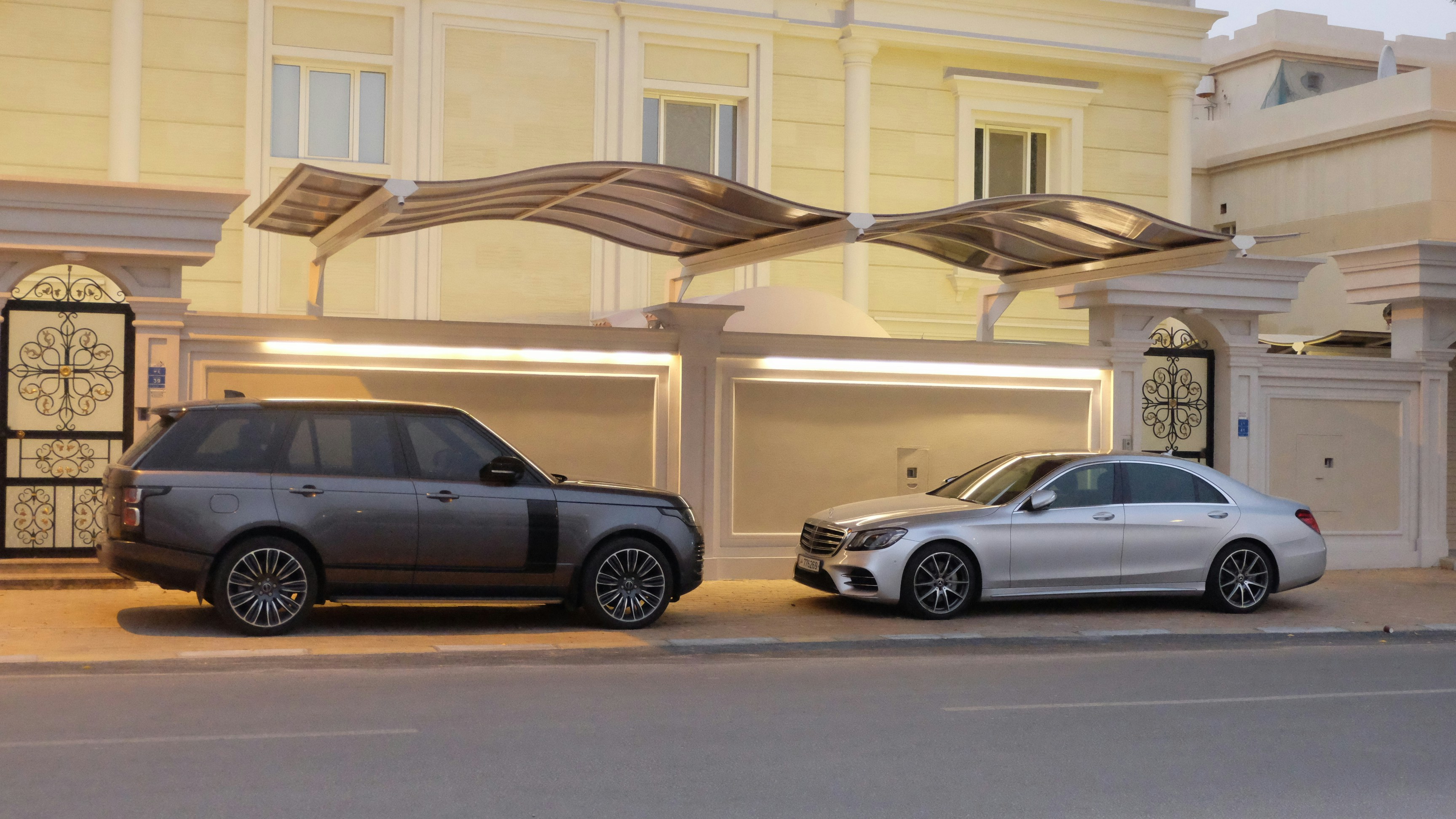 Two luxury cars parked side by side beneath a modern canopy in an upscale neighborhood, showcasing architectural elegance and automotive design.