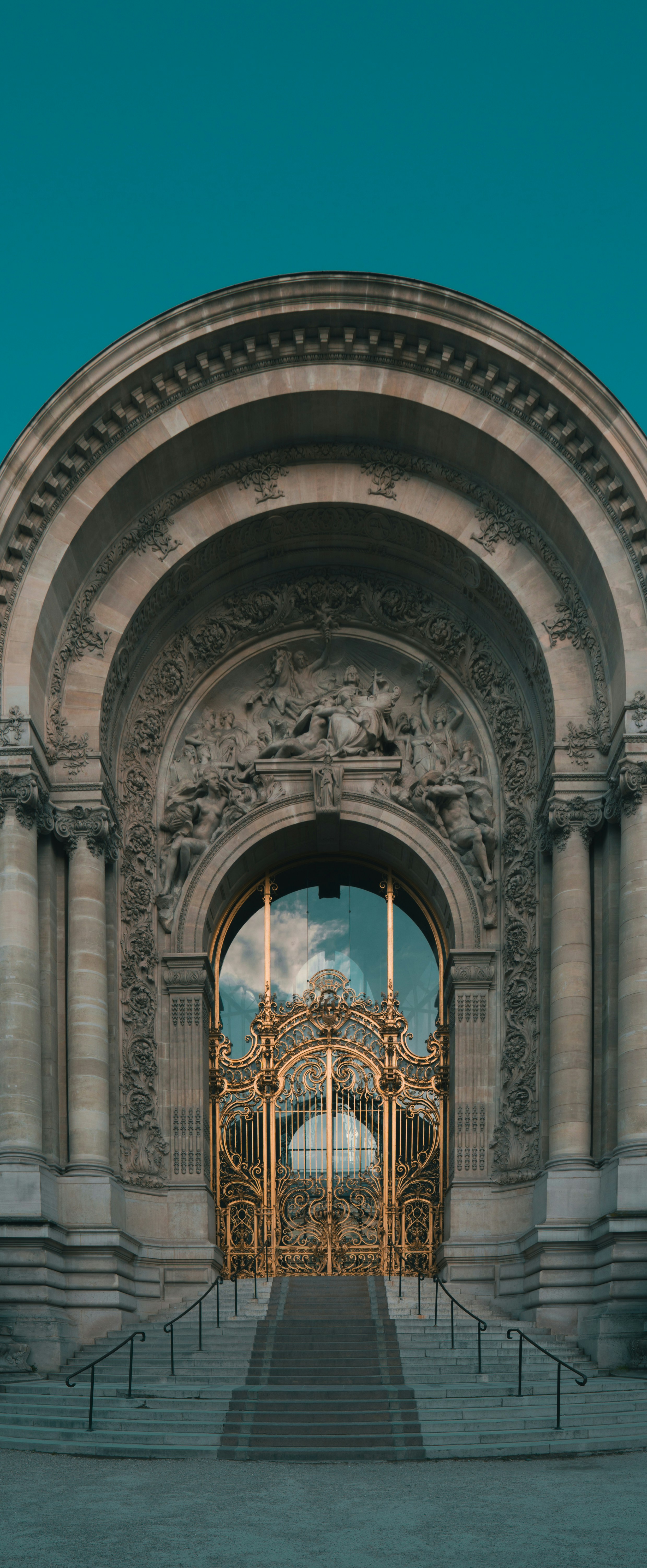 Intricately designed golden gates framed by ornate stone architecture, reflecting the sky in the background.