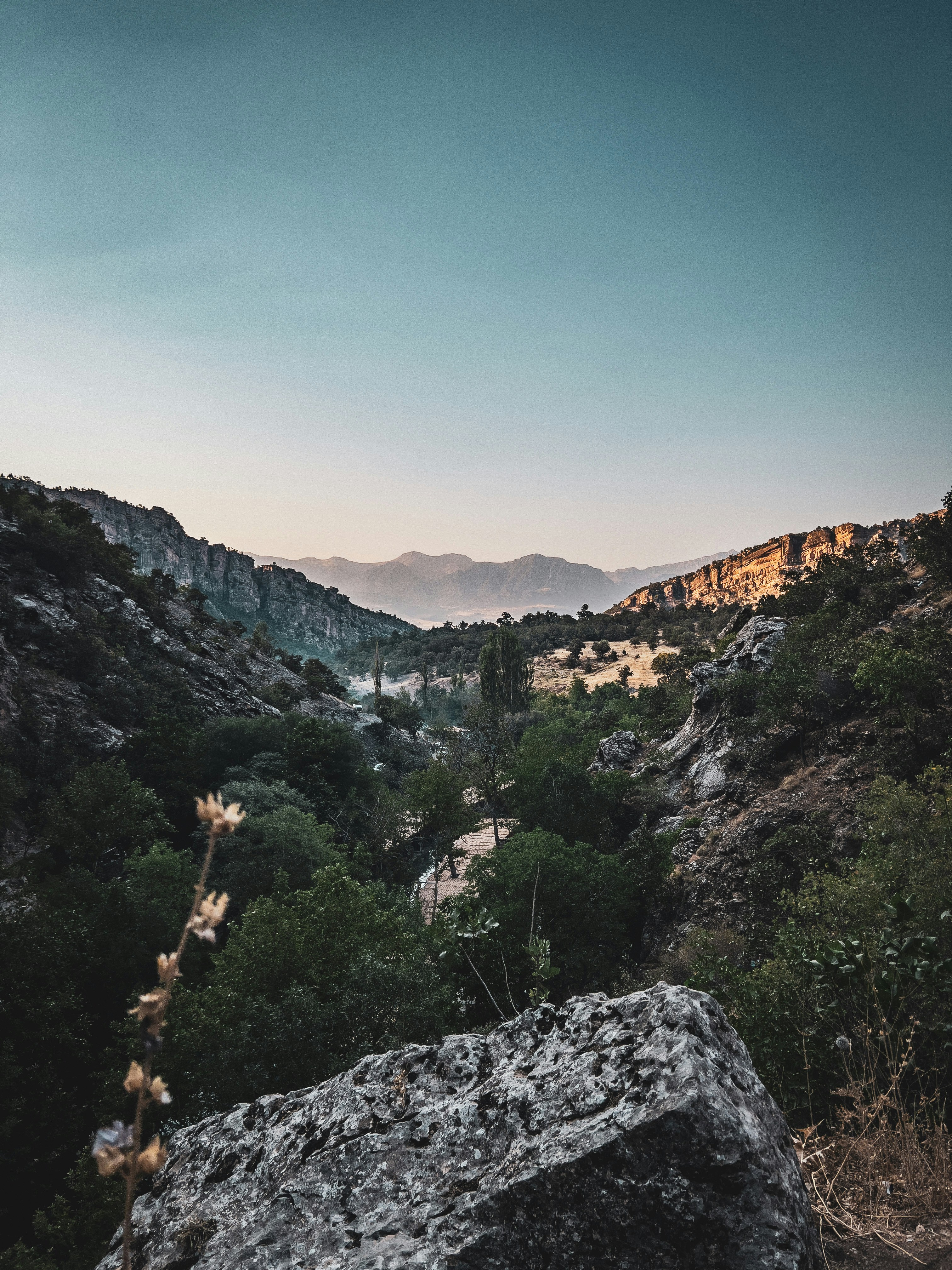 Serene valley landscape bathed in warm evening light, framed by rocky outcrops and lush vegetation. The distant mountains create a tranquil backdrop.
