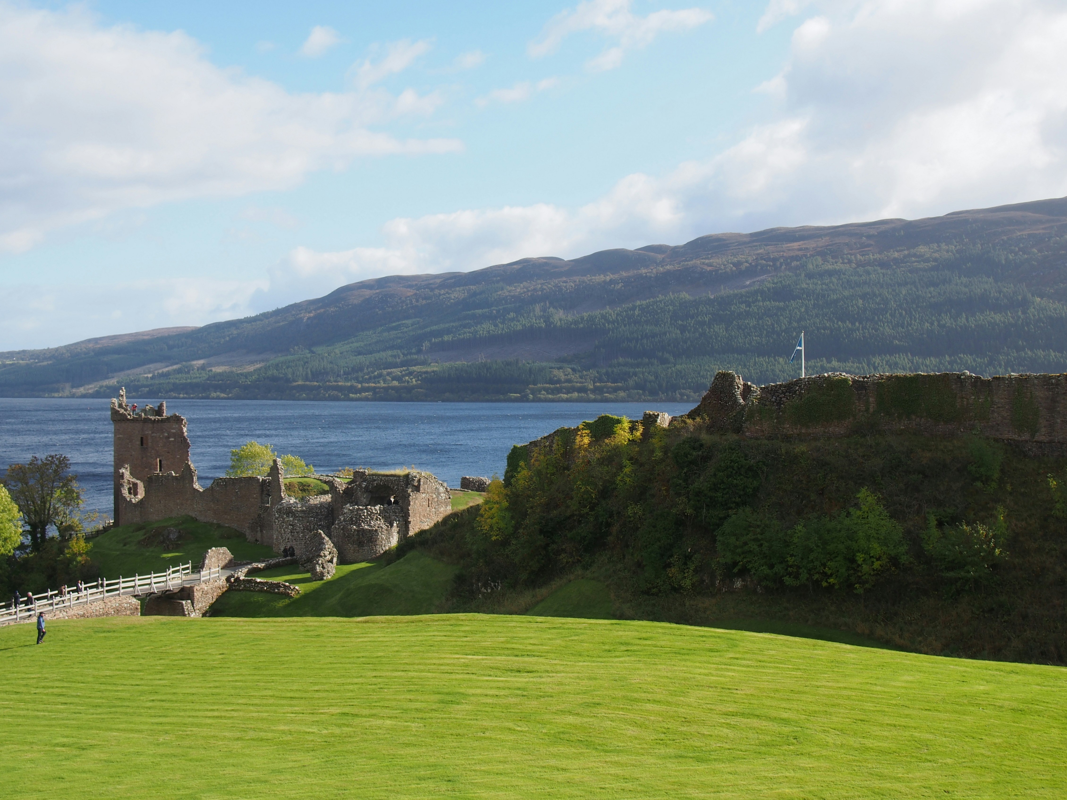 Ancient castle ruins overlook a tranquil loch with distant rolling hills under a partly cloudy sky.