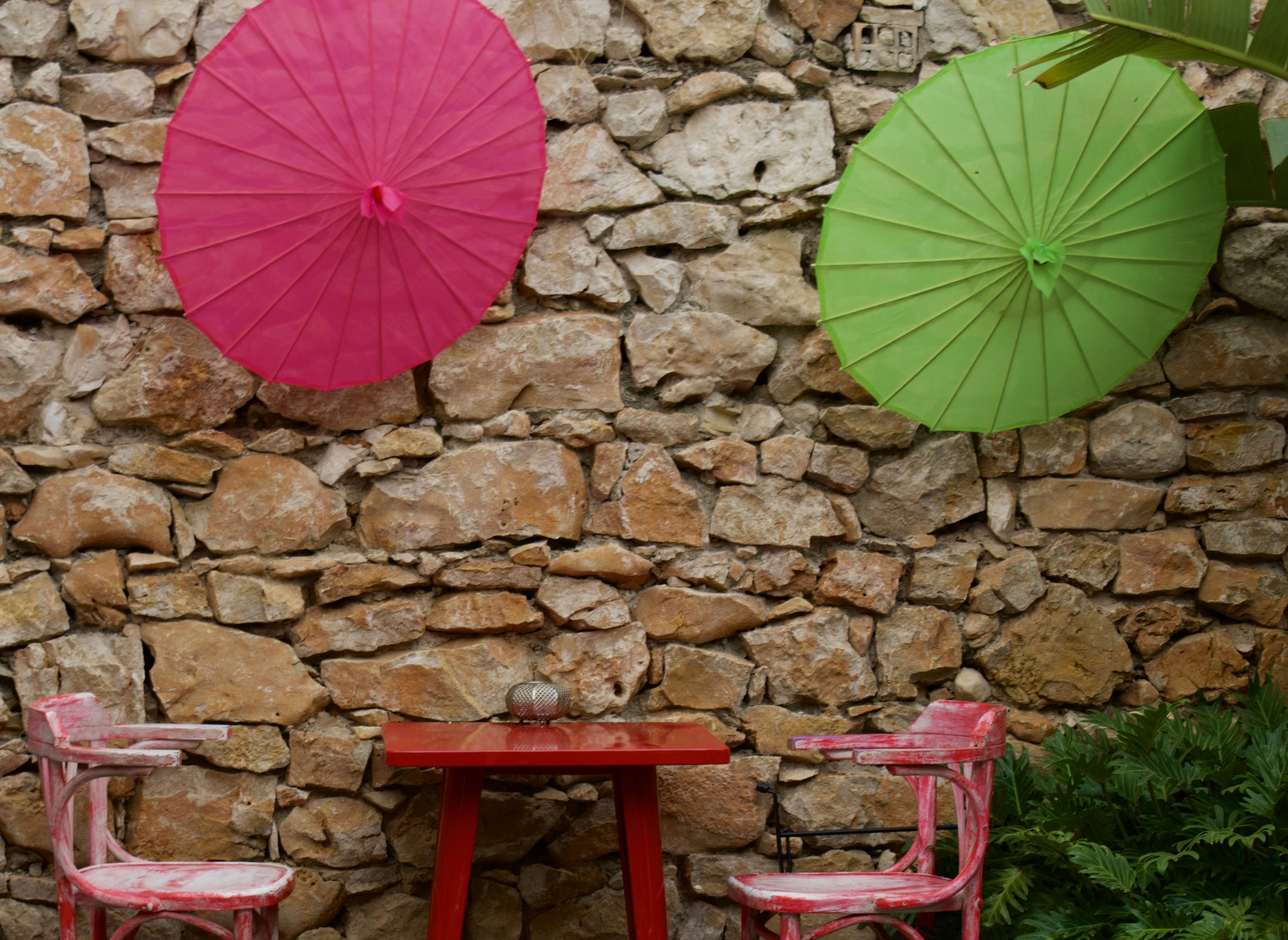 Crimson Table, Emerald Umbrellas: A Quiet Corner Against a Stone Wall