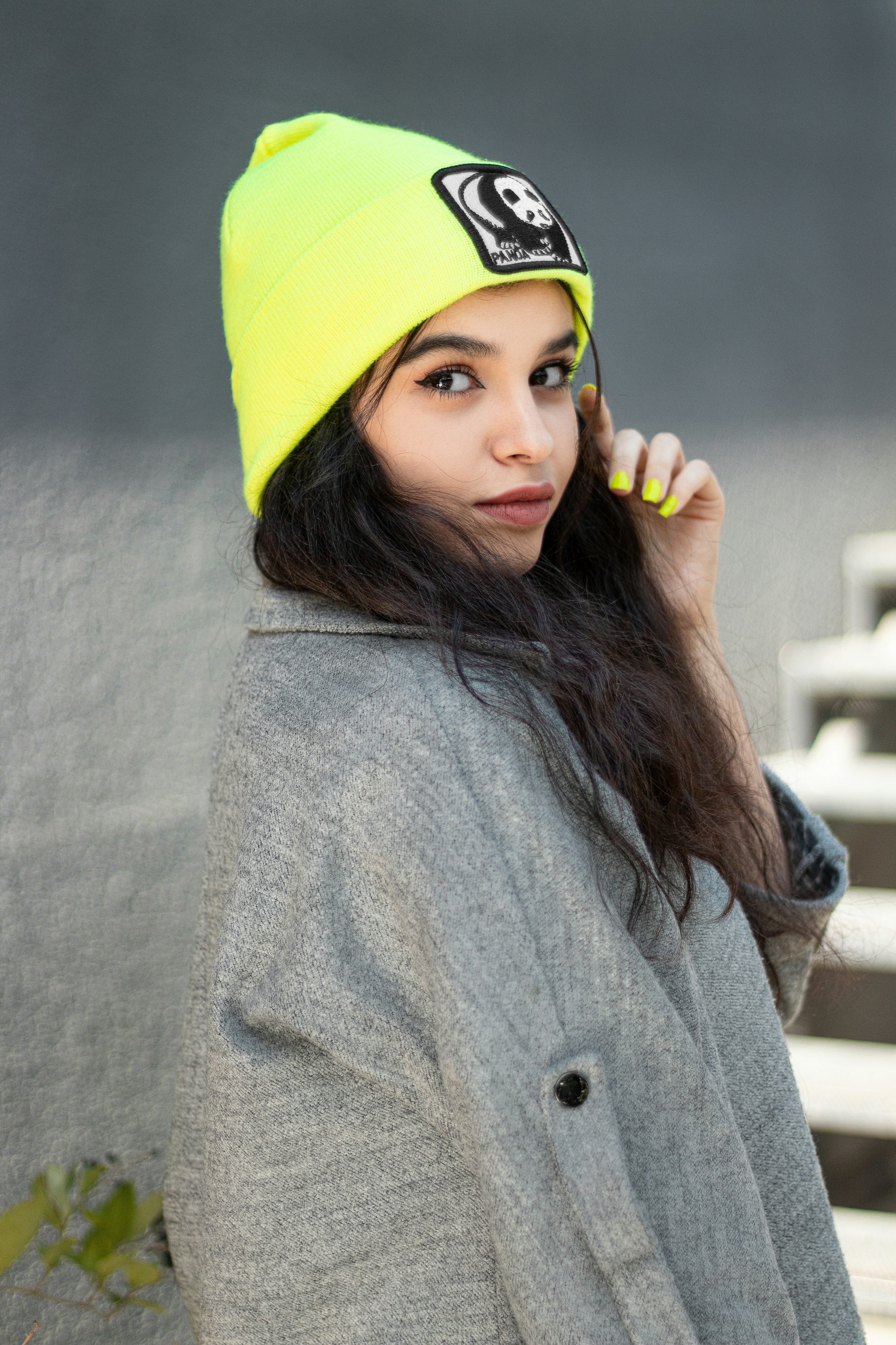 Young woman in a vibrant yellow beanie and gray coat poses against a muted background, exuding a modern urban style.