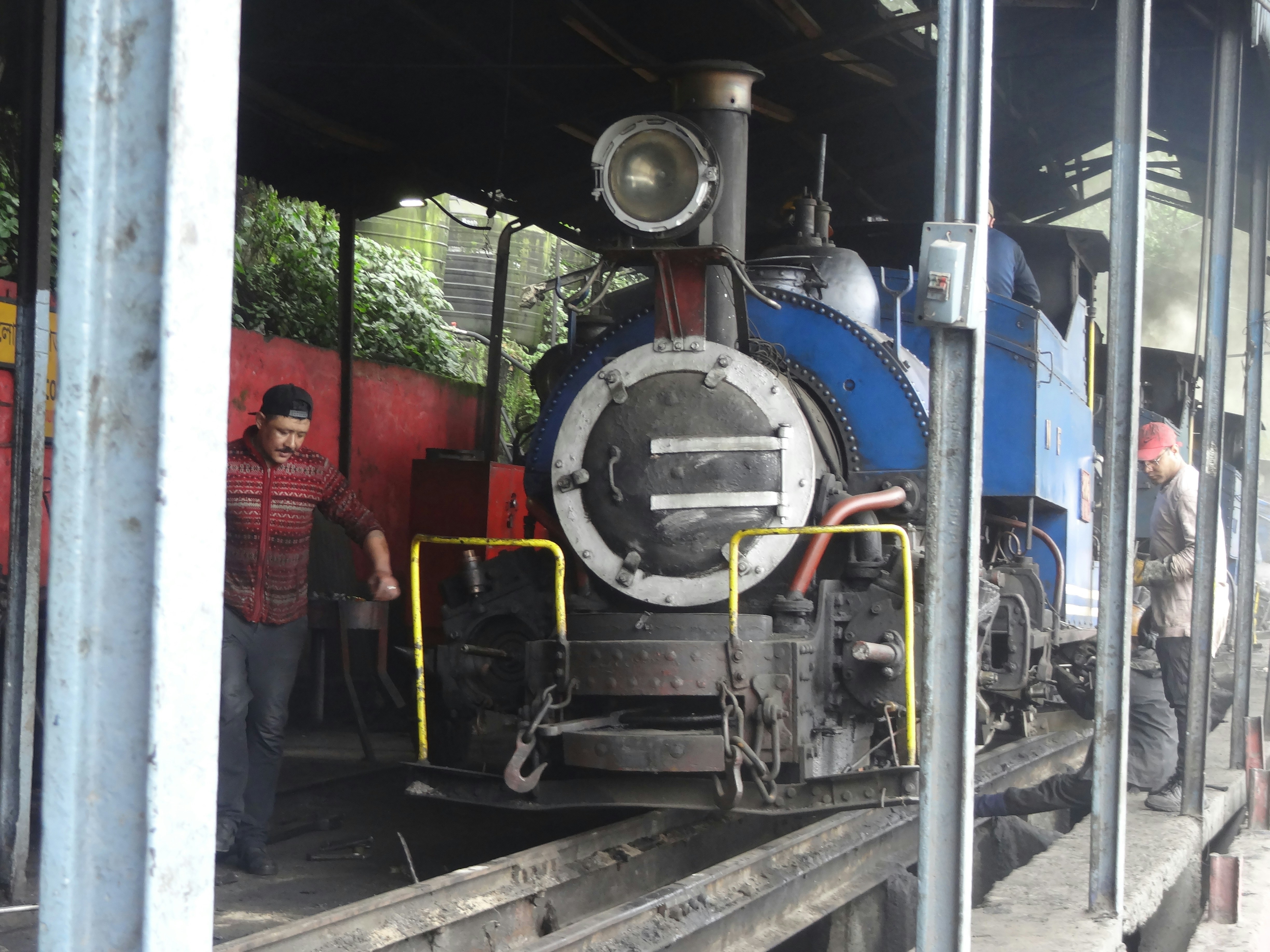Blue steam locomotive being serviced in a workshop, with workers attending to it amidst industrial surroundings.