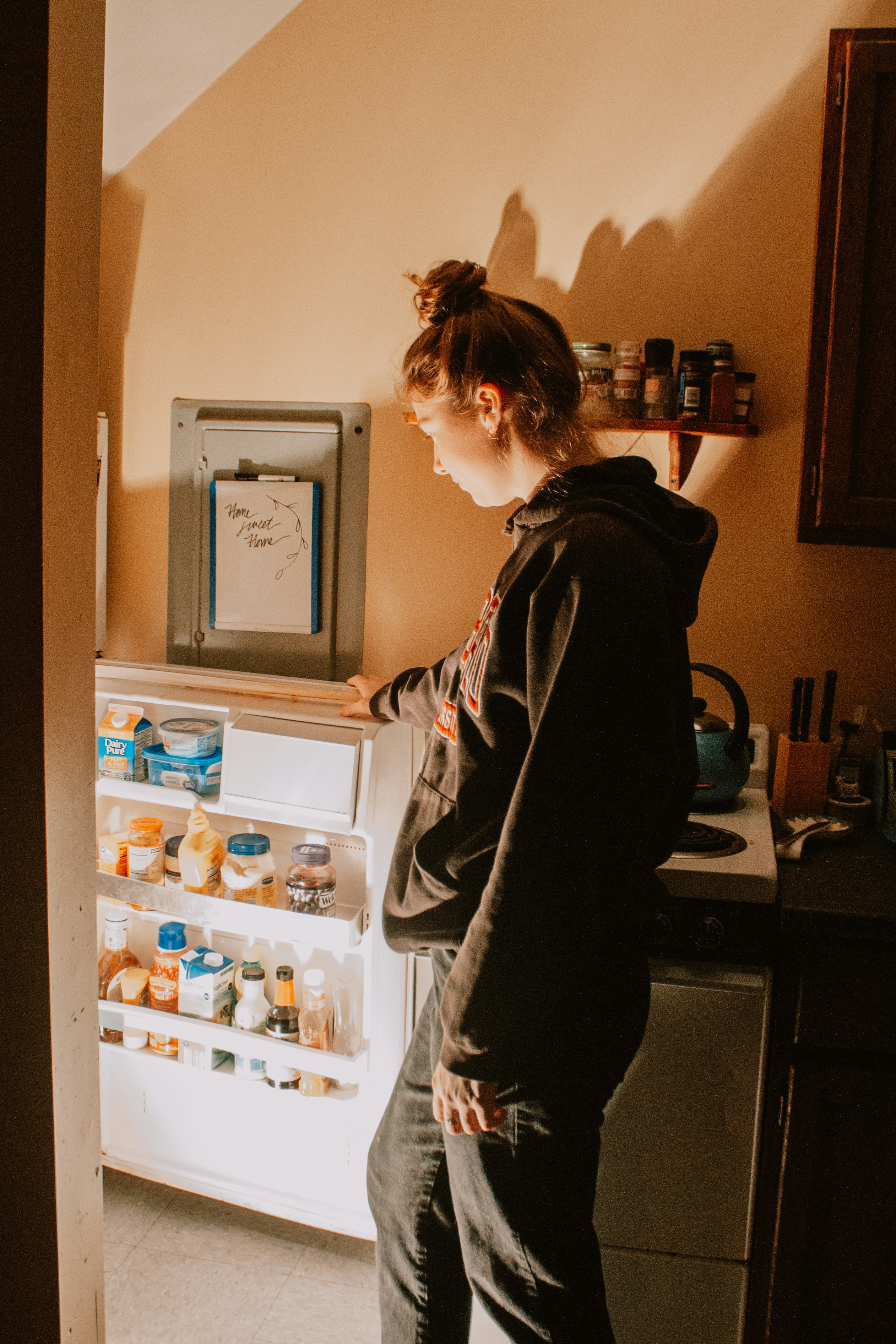 Young person pondering choices while gazing into an open refrigerator filled with various drinks and condiments.