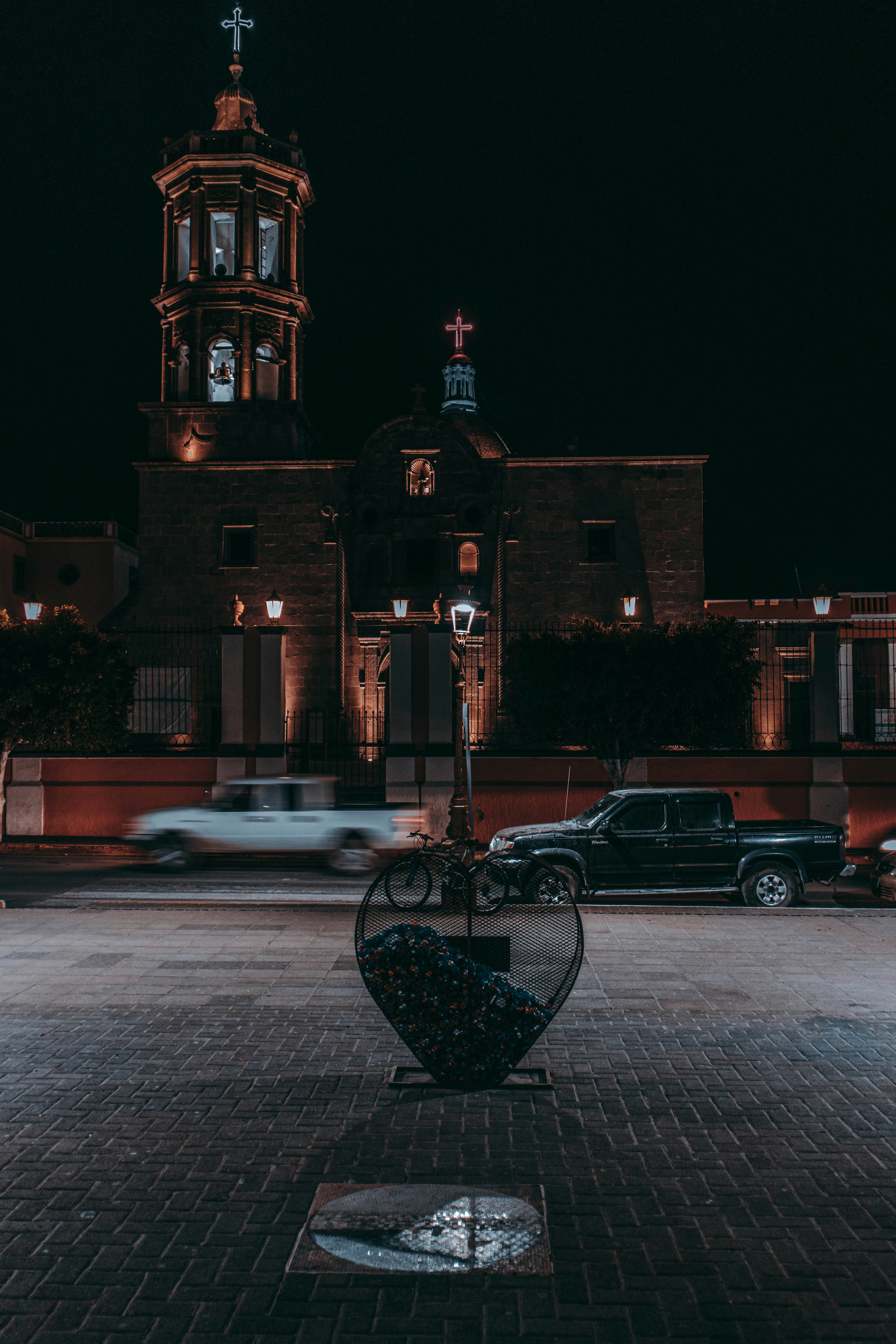 A heart-shaped metal planter anchors the foreground of a dimly lit plaza, its silhouette illuminated by street lamps, while a brick church rises in the background against the night sky.