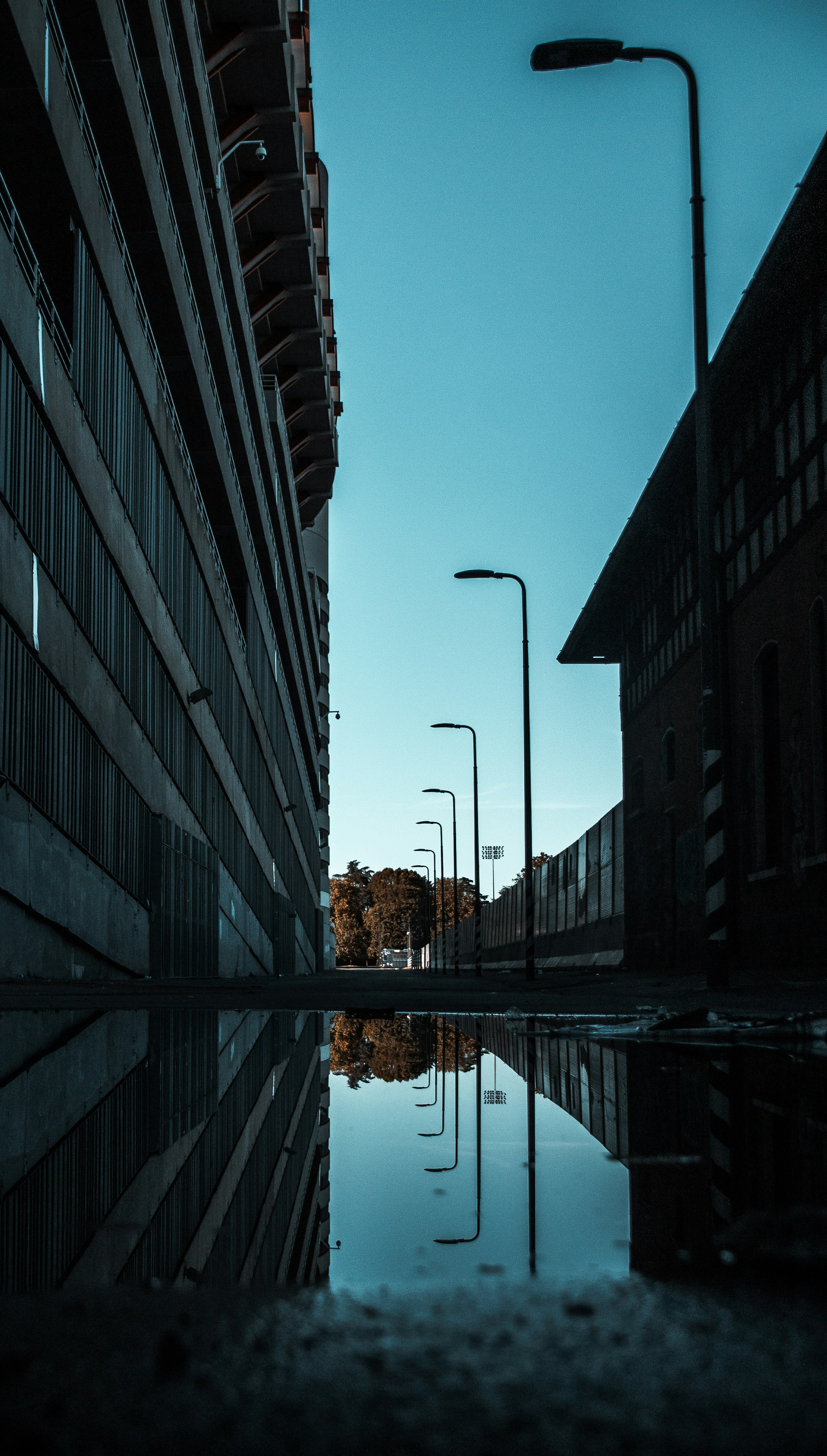 A quiet urban alleyway with buildings on either side, featuring a reflective puddle that captures the structures and streetlights above.