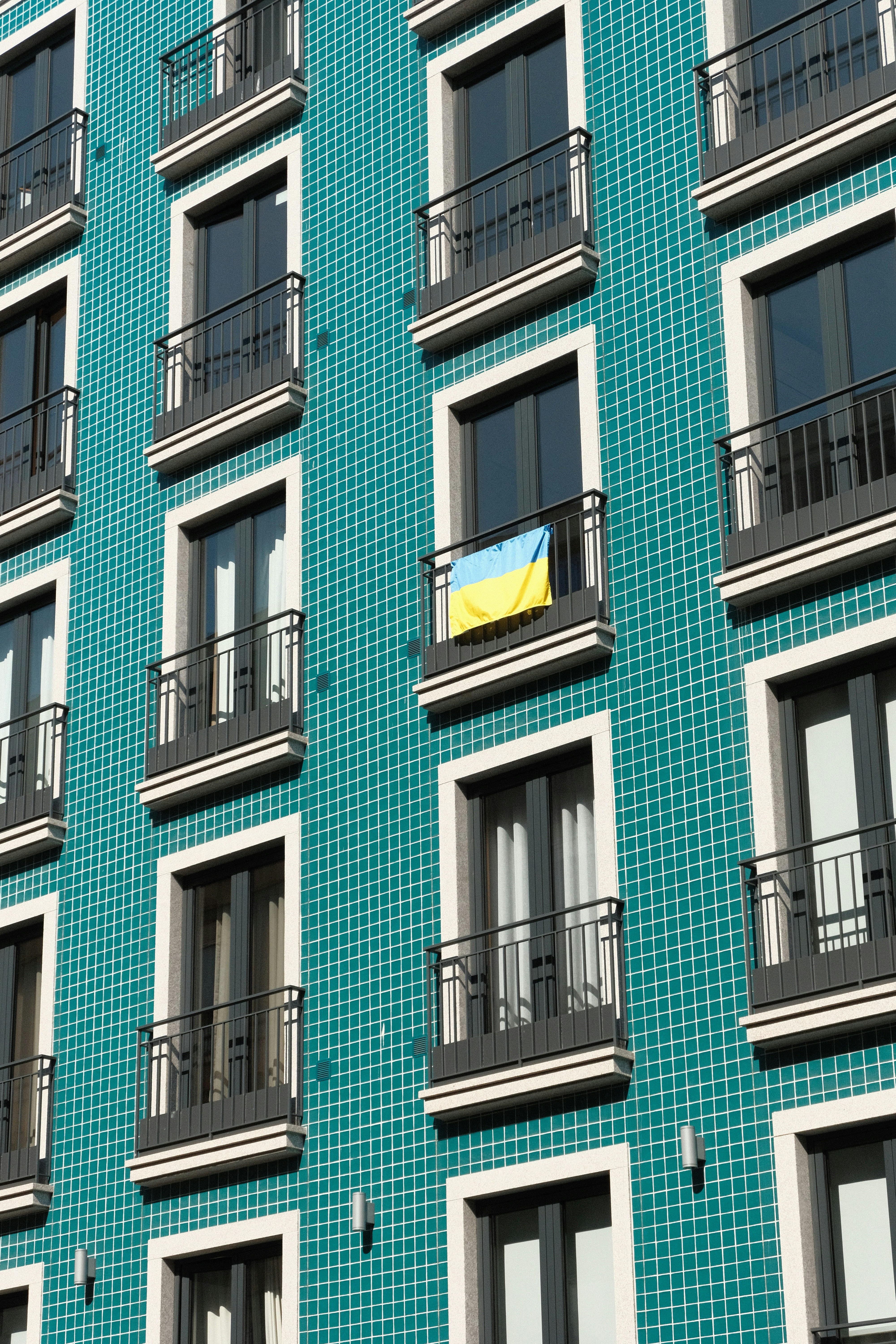 Photograph of a teal-tiled apartment building facade with uniform balconies, featuring a blue-and-yellow flag on a central balcony.