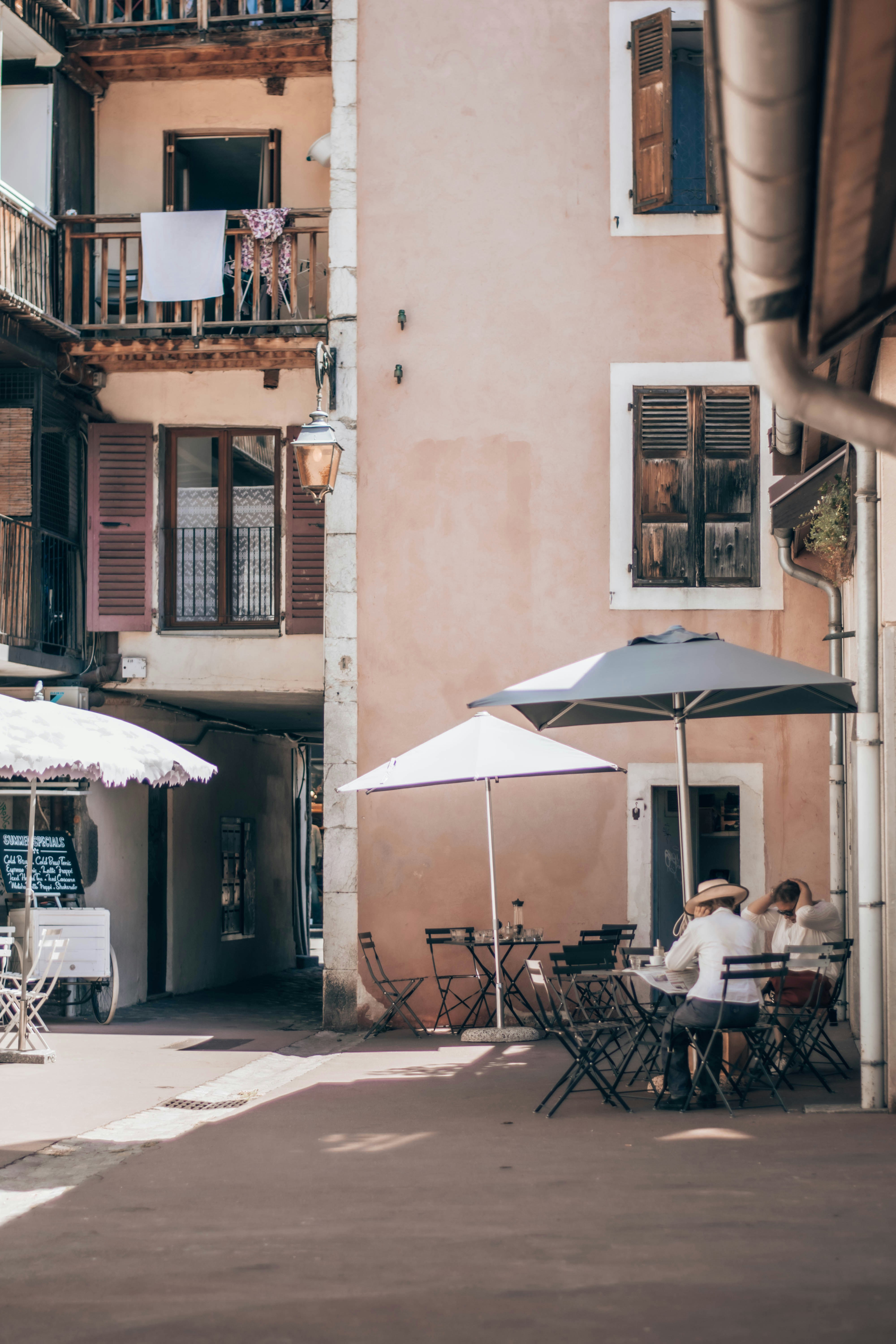 Outdoor café scene featuring patrons under large umbrellas in a quaint alley, surrounded by pastel-colored buildings and rustic shutters.