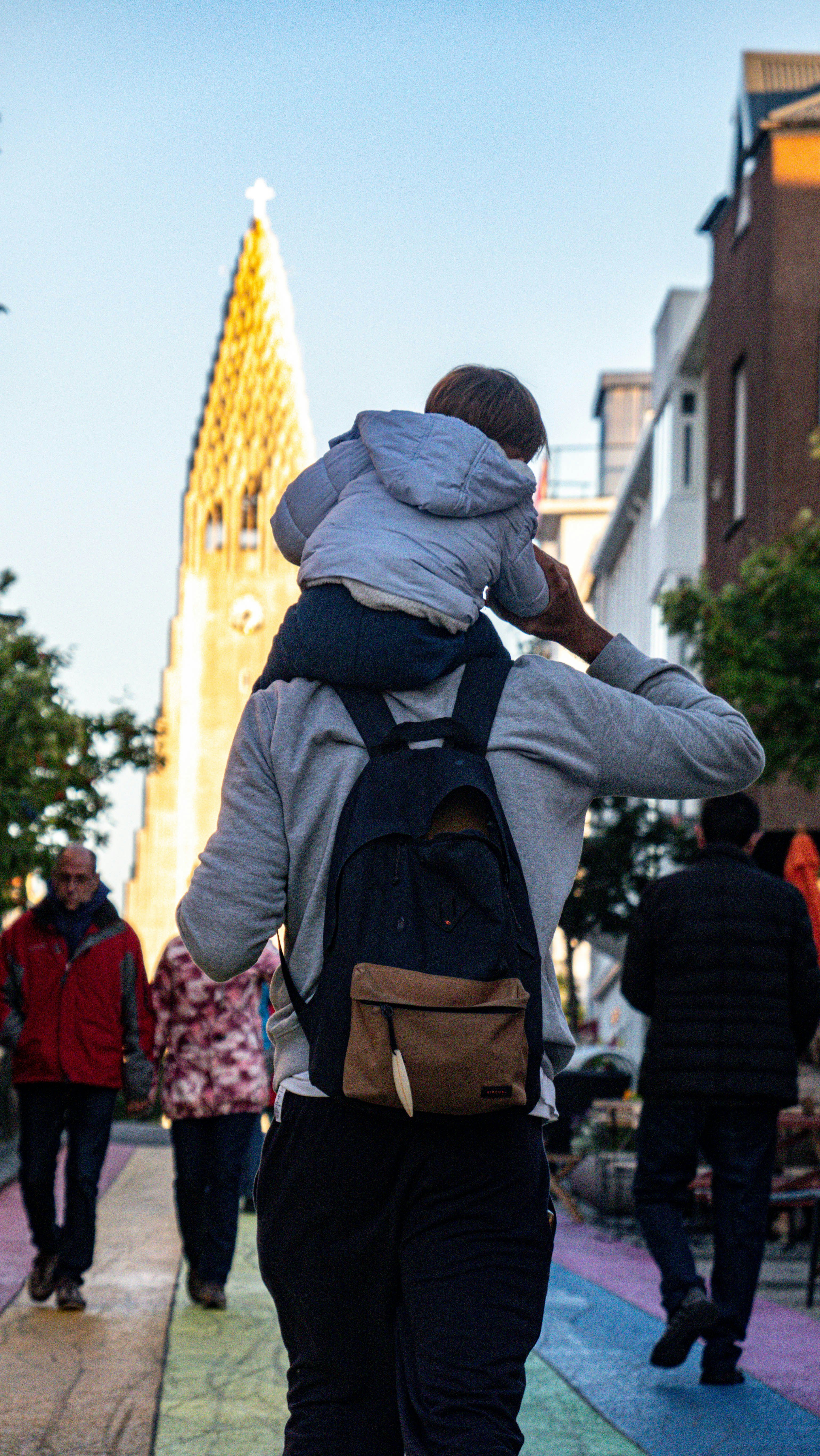 A man carries a child on his shoulders as they walk along a rainbow-colored pedestrian street toward a church spire. Passersby and urban buildings frame the lively scene.