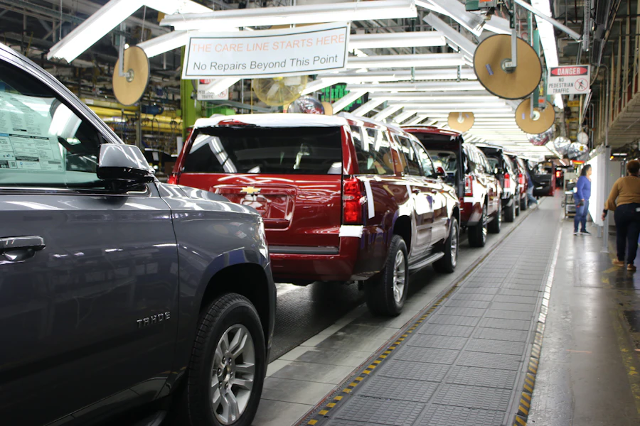 Row of vehicles on an automotive assembly line under bright factory lighting. The photo emphasizes industrial throughput and the scale of vehicle production.
