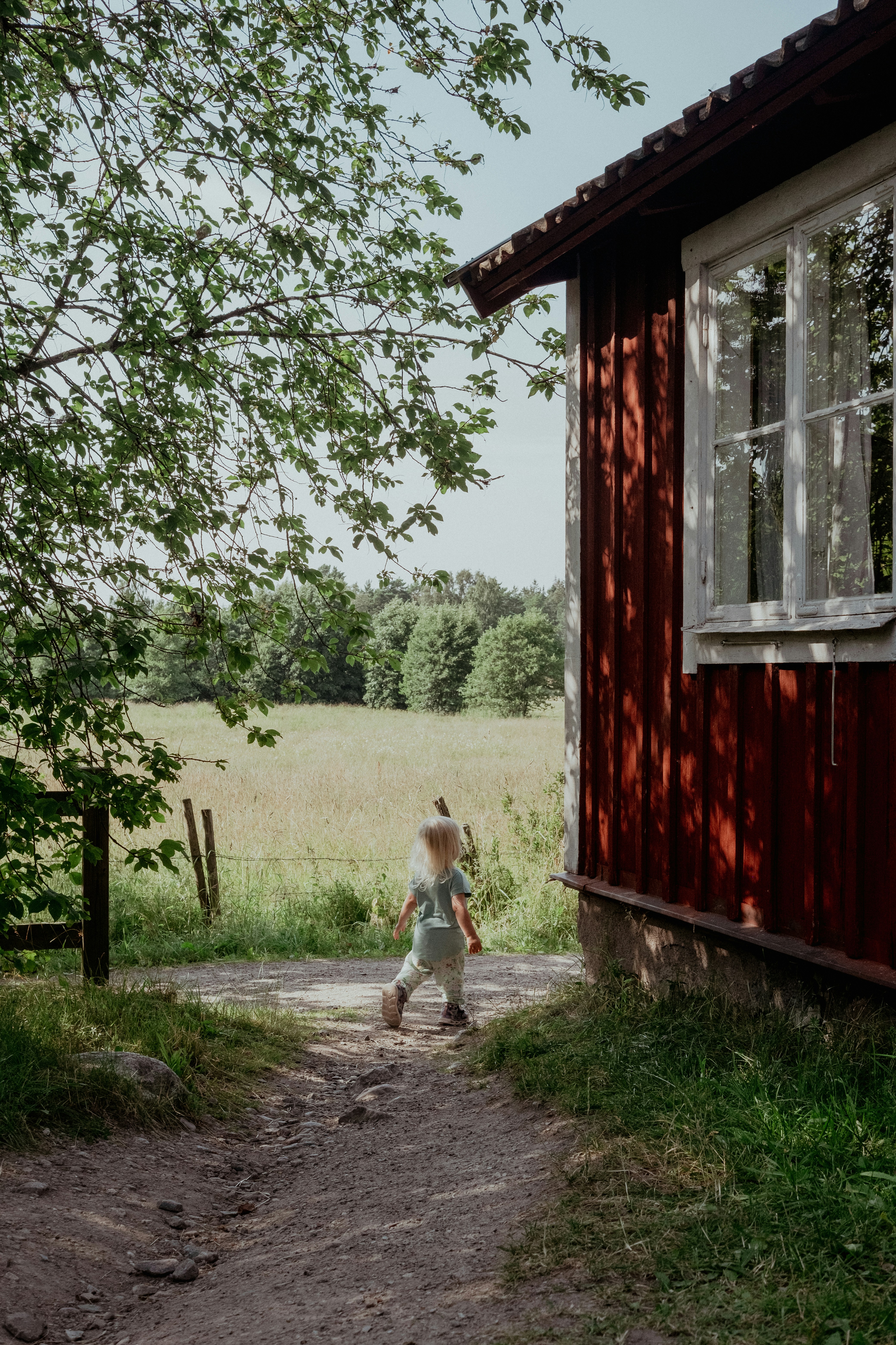 Child walking along a dirt path beside a rustic red cottage, framed by lush greenery and open fields.