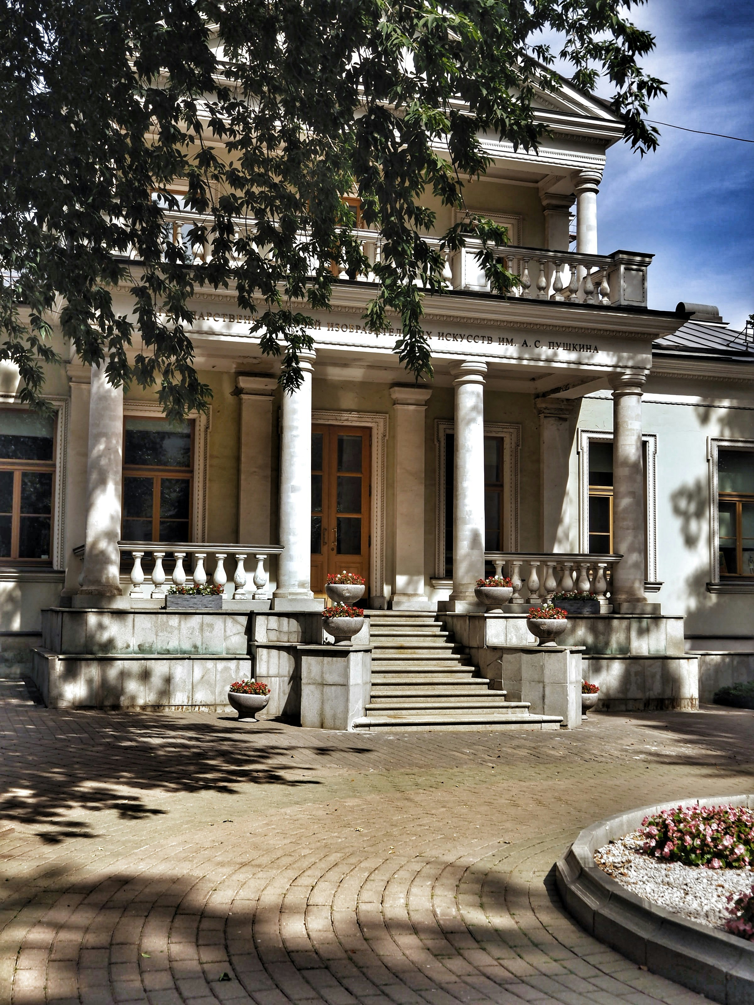 Grand neoclassical mansion with ornate columns and a staircase, surrounded by lush greenery and flower pots. The architectural details reflect a blend of history and sophistication.