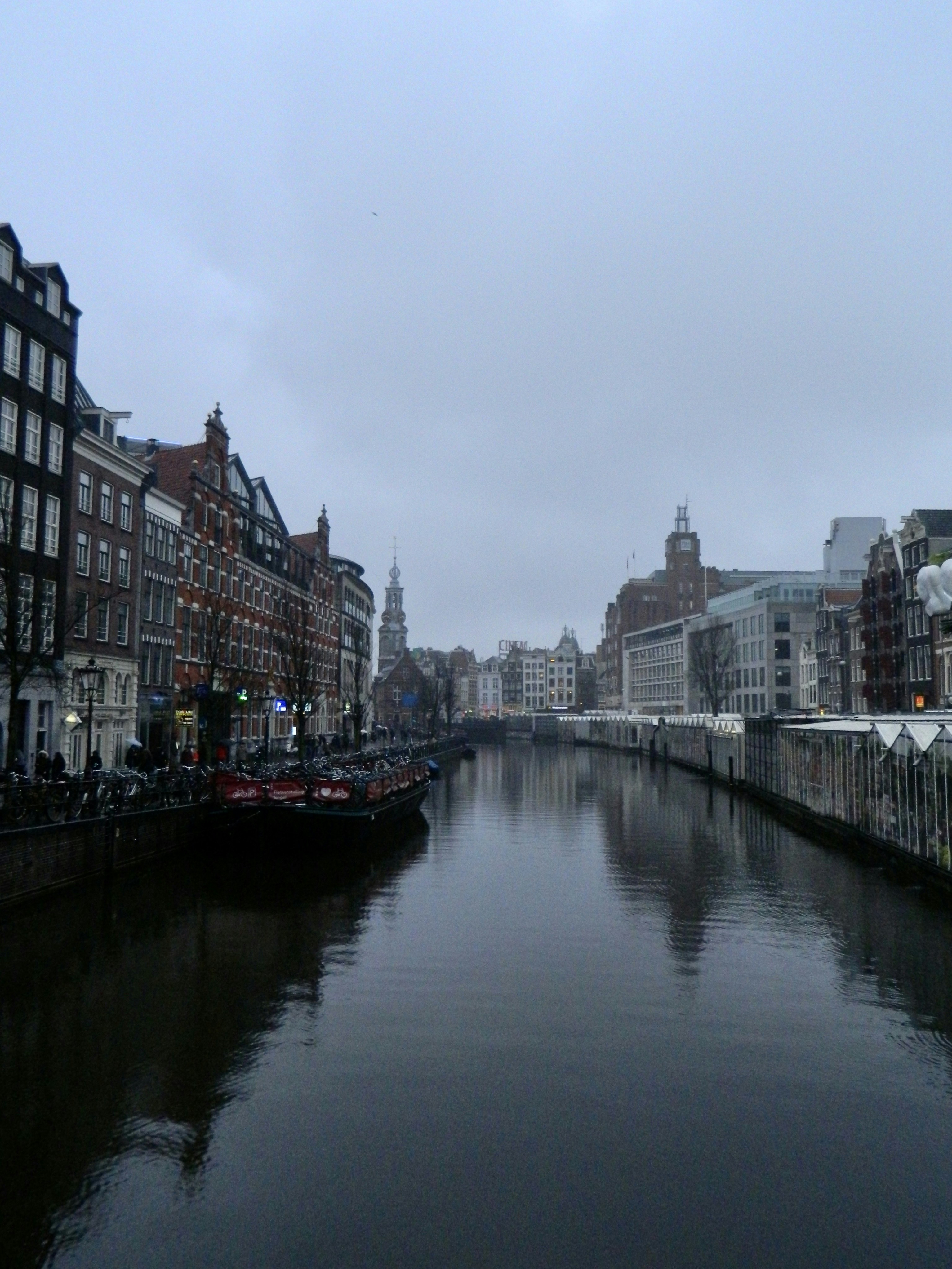 Canal lined with historic buildings and moored boats under a cloudy sky. The tranquil water reflects the architecture along the banks.