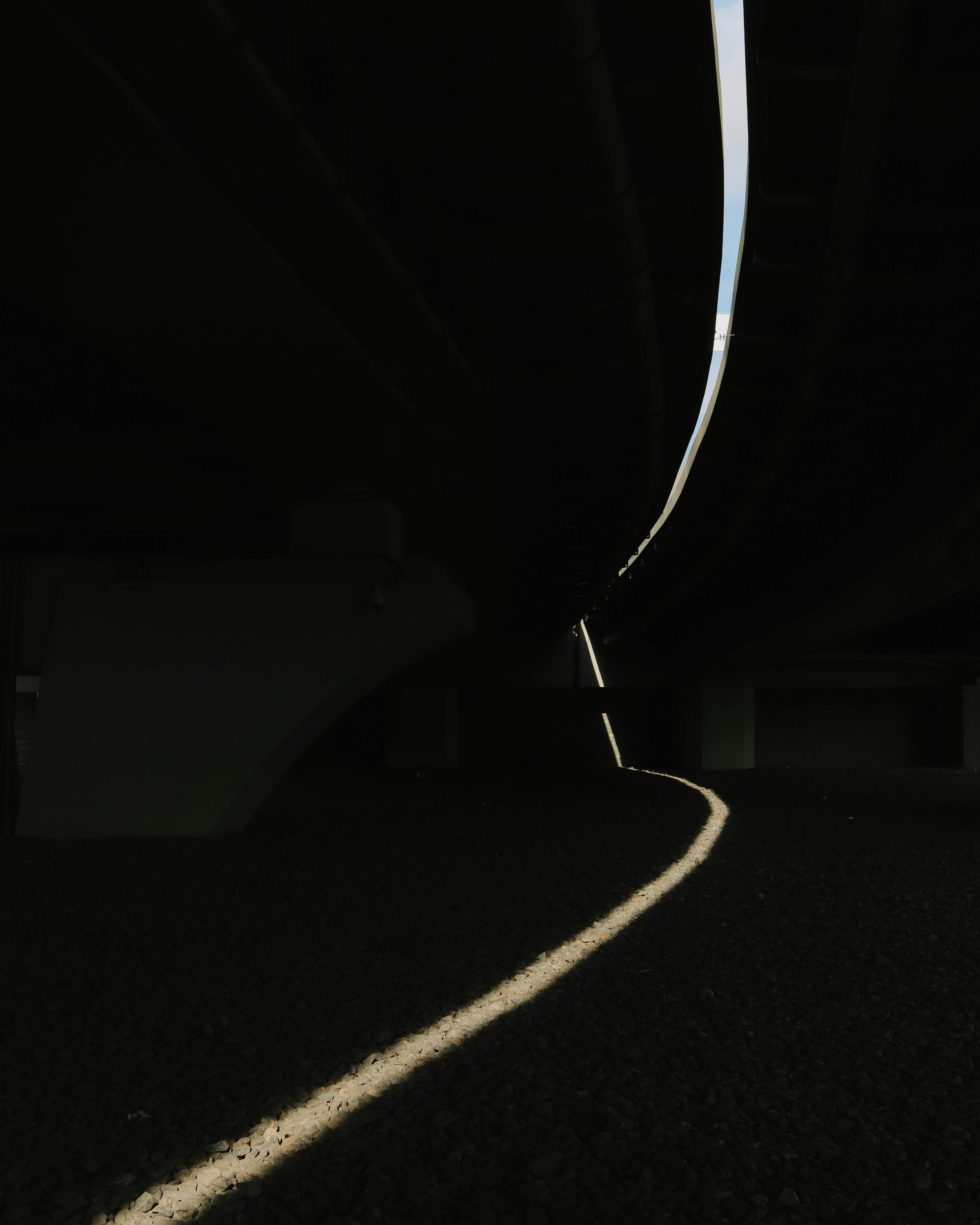 A narrow beam of light cuts through the darkness under a concrete structure, creating a striking contrast with the surrounding shadows.