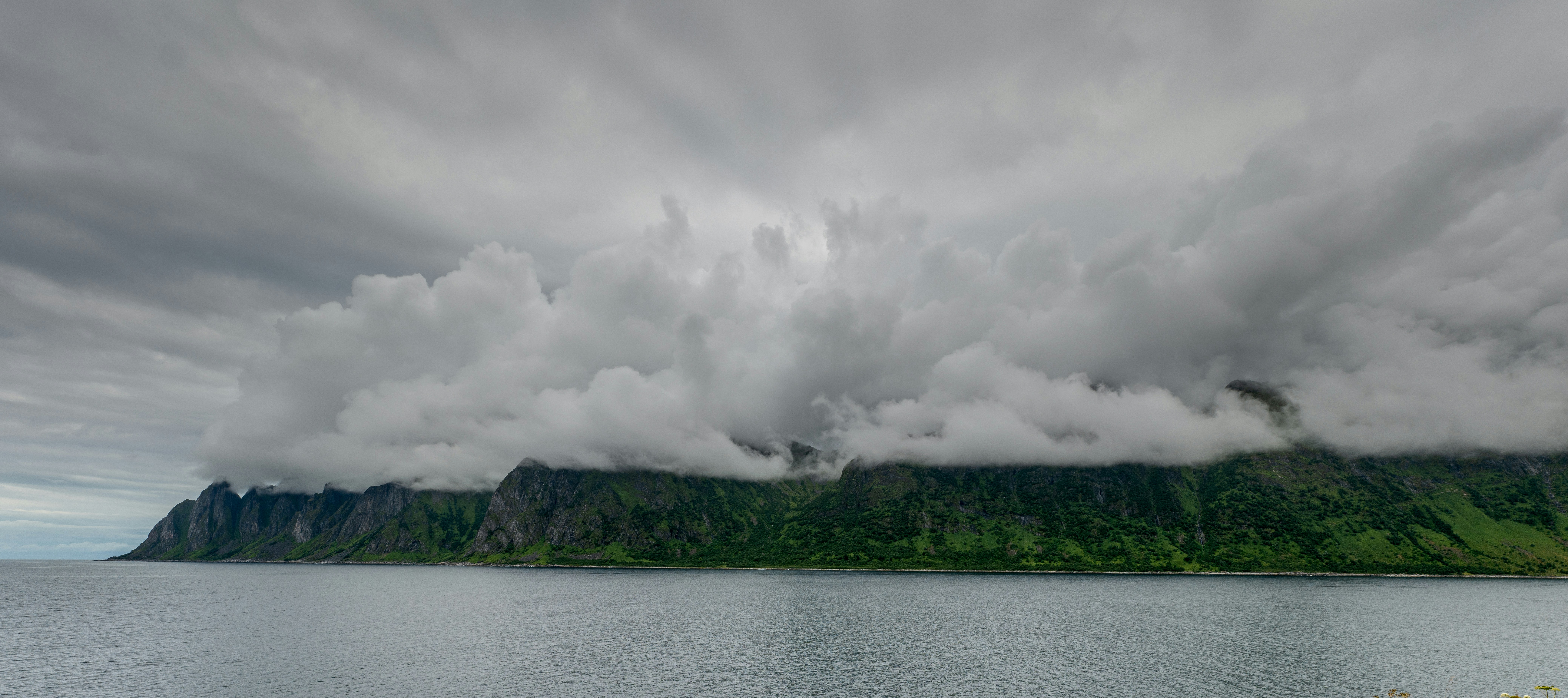 Dramatic clouds hover over a lush green mountain range by a calm sea under a gray sky.