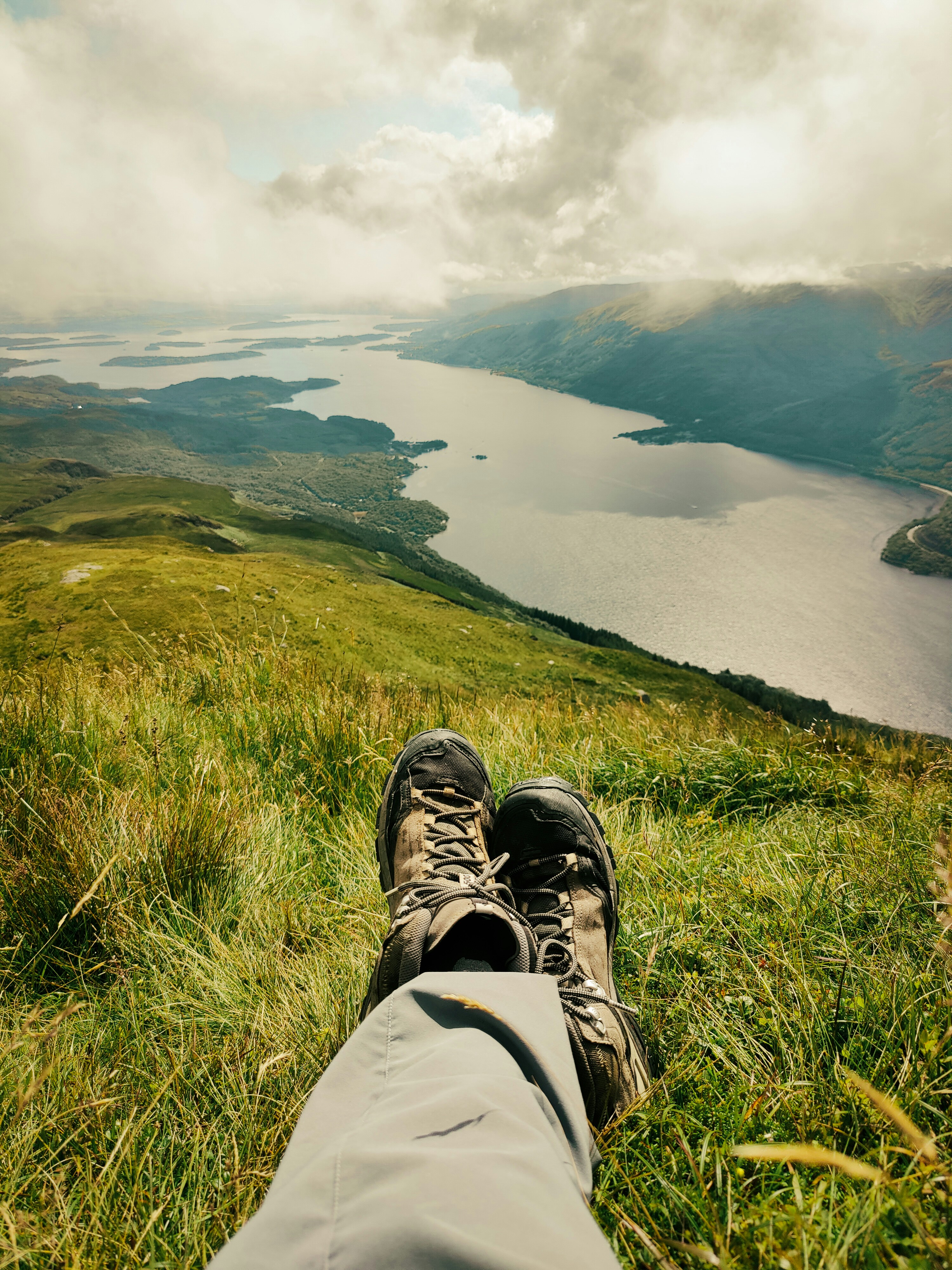 Worn hiking boots resting on grassy hillside overlooking Loch Lomond under a cloudy sky.