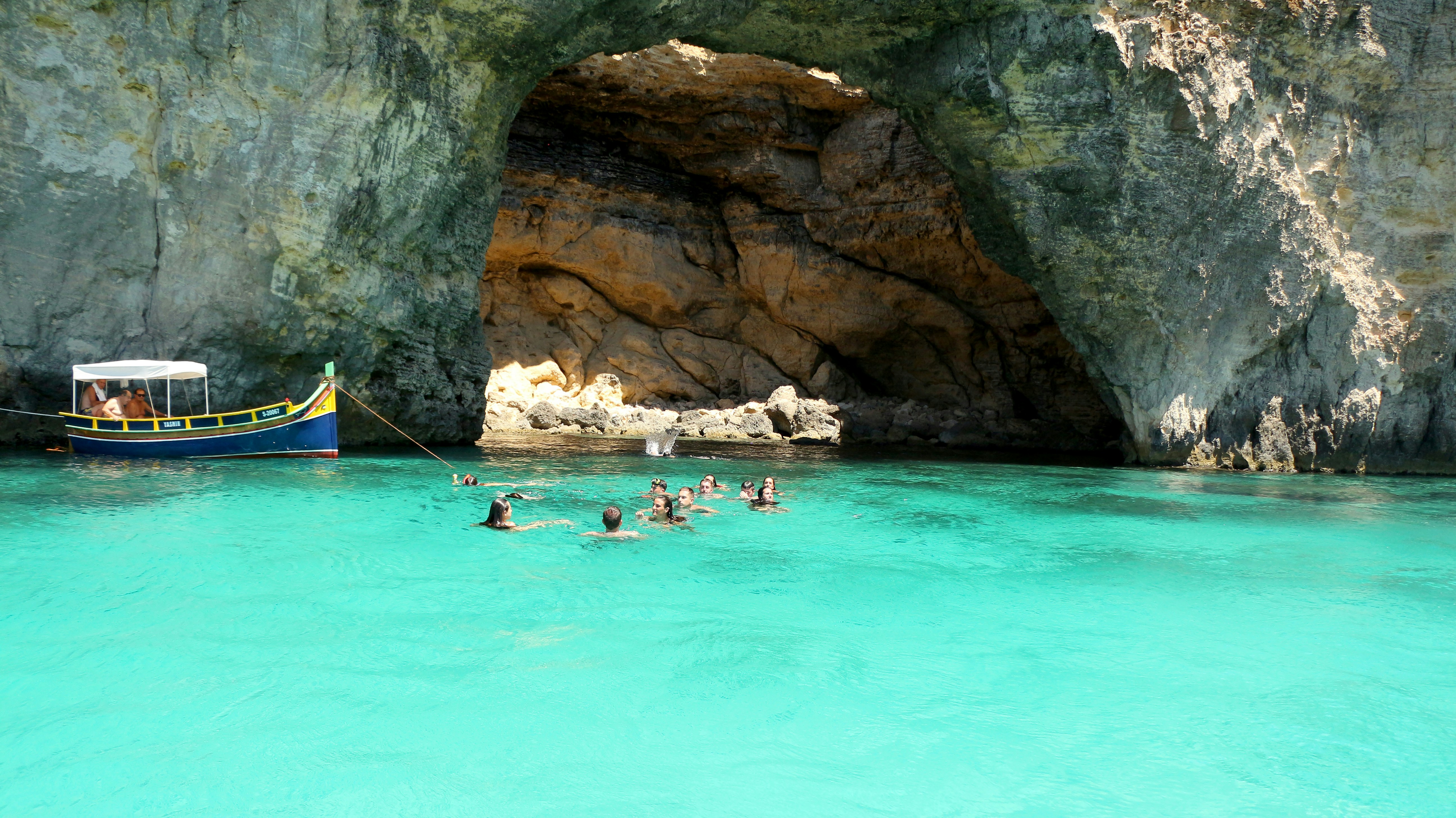 Swimmers enjoying the vibrant turquoise waters near a rocky cave, with a traditional boat anchored nearby.