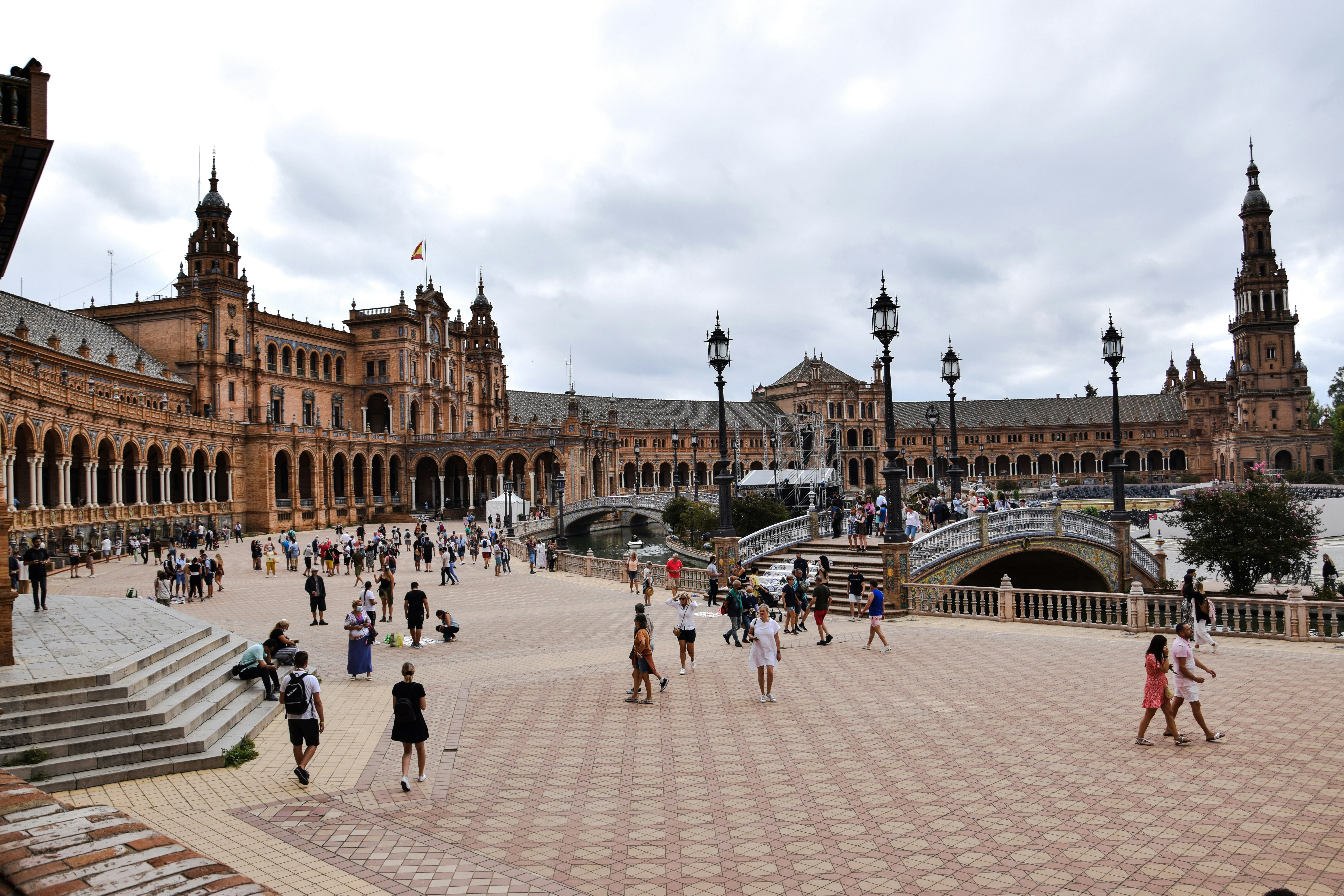 Visitors stroll through the expansive Plaza de España under a cloudy sky, surrounded by ornate architecture and bridges.