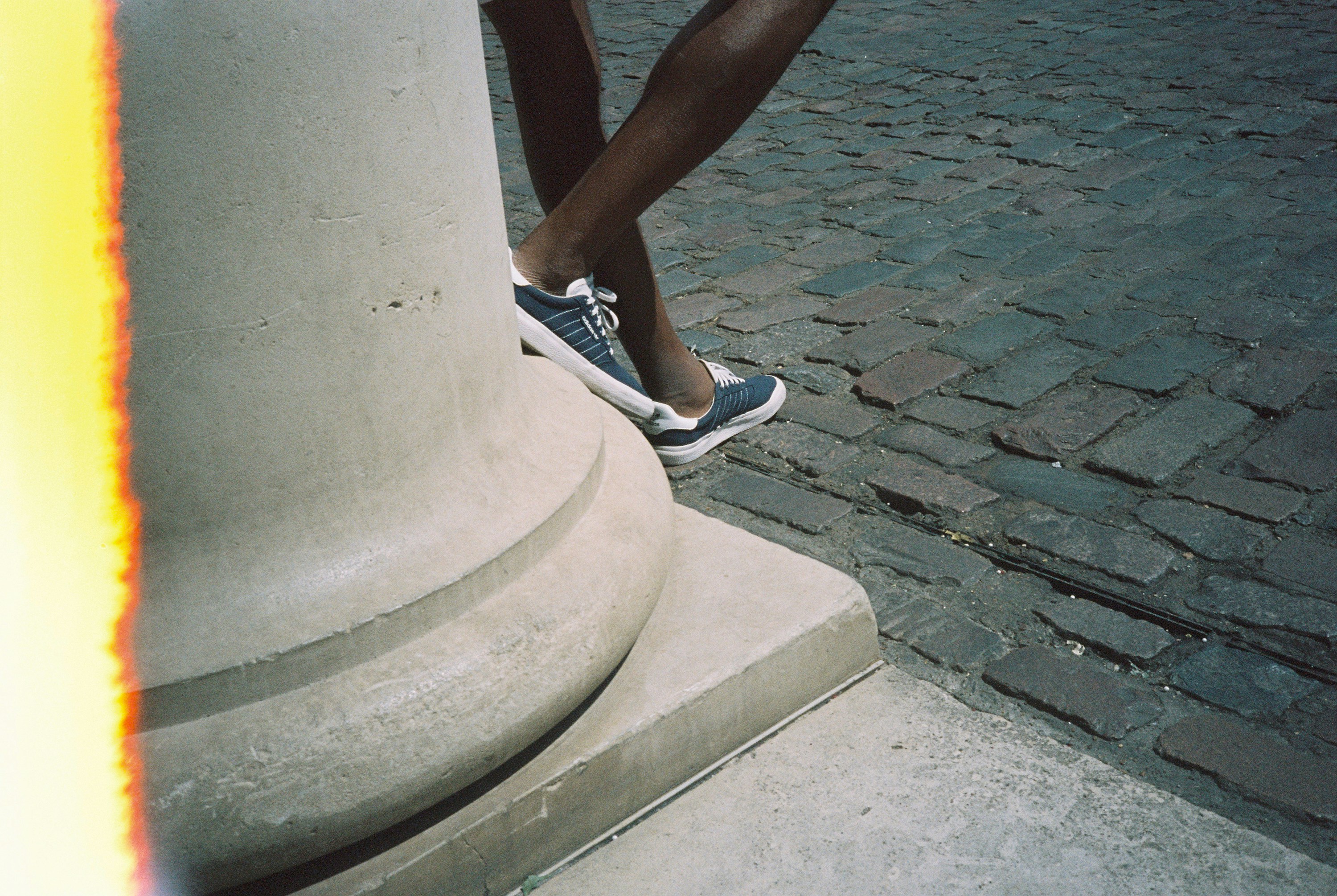 Bare legs and blue Adidas training shoes or sneakers. A man leaning against a stone pillar. Shot on 35mm film, there is a light leak on the left hand side.