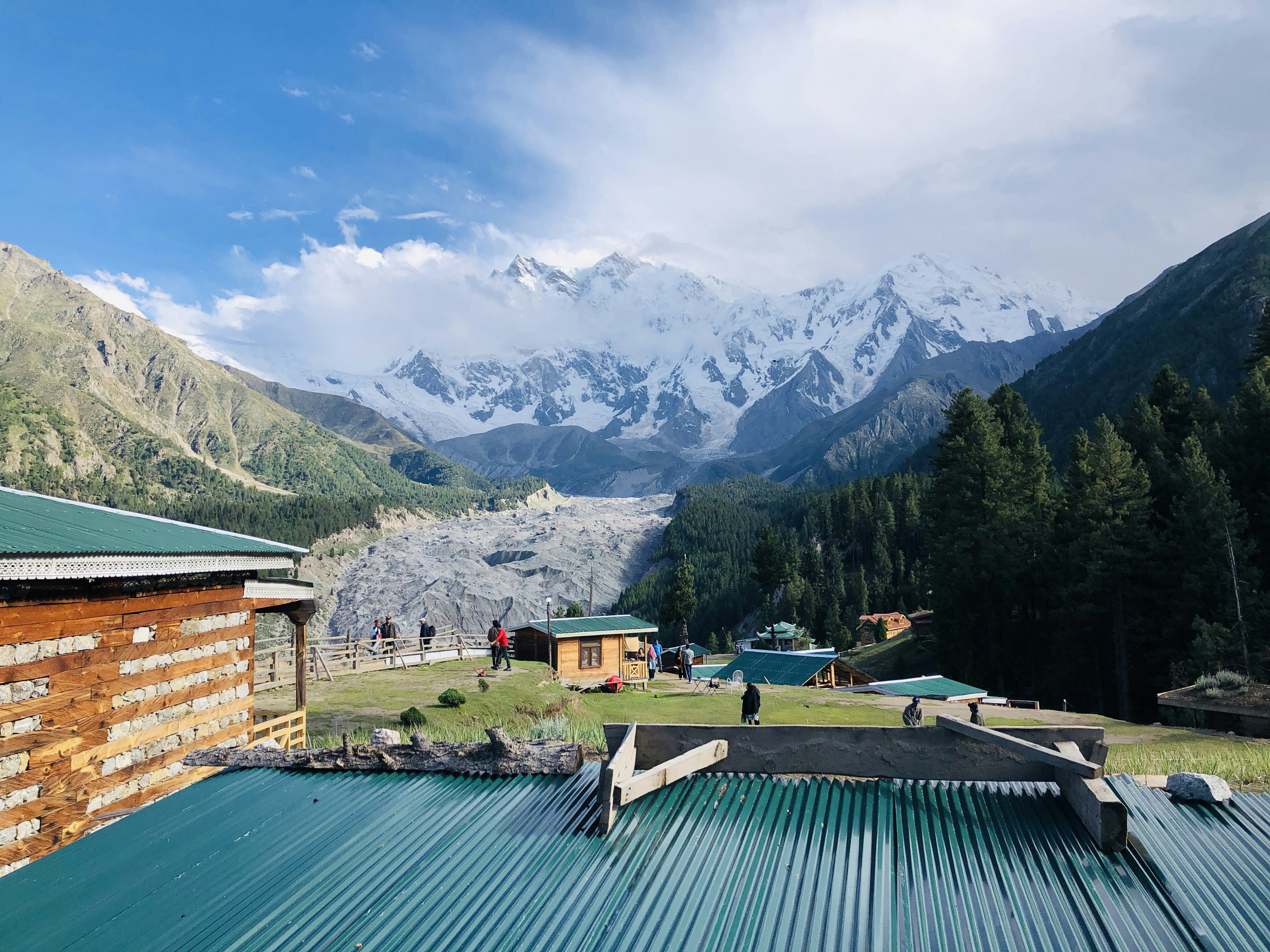 Rustic cabins nestled in a forest with towering snow-capped mountains under a bright blue sky.