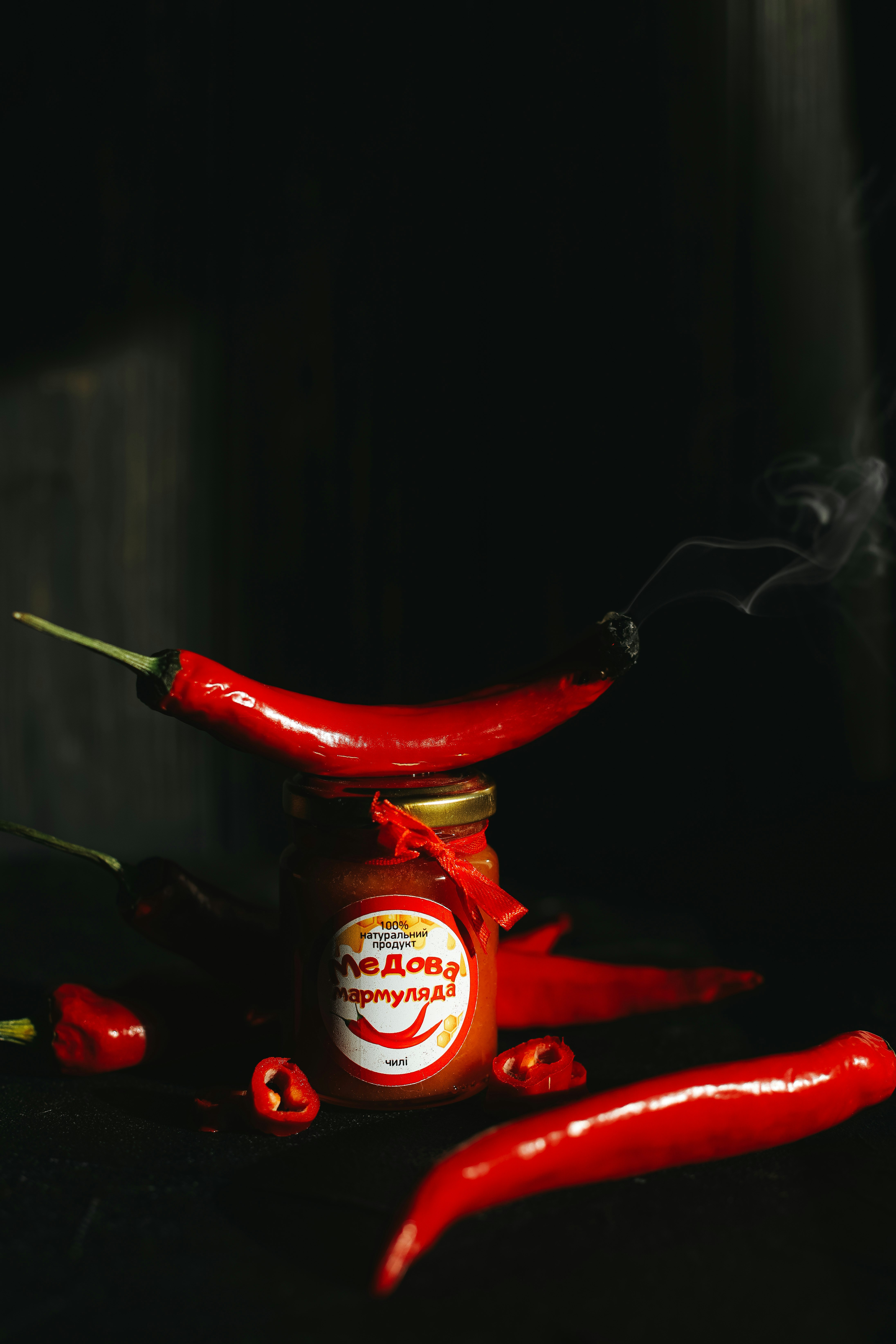 A jar of spicy red chili paste surrounded by vibrant red chilies, with smoke rising from the top. The dark background enhances the vivid colors and textures.