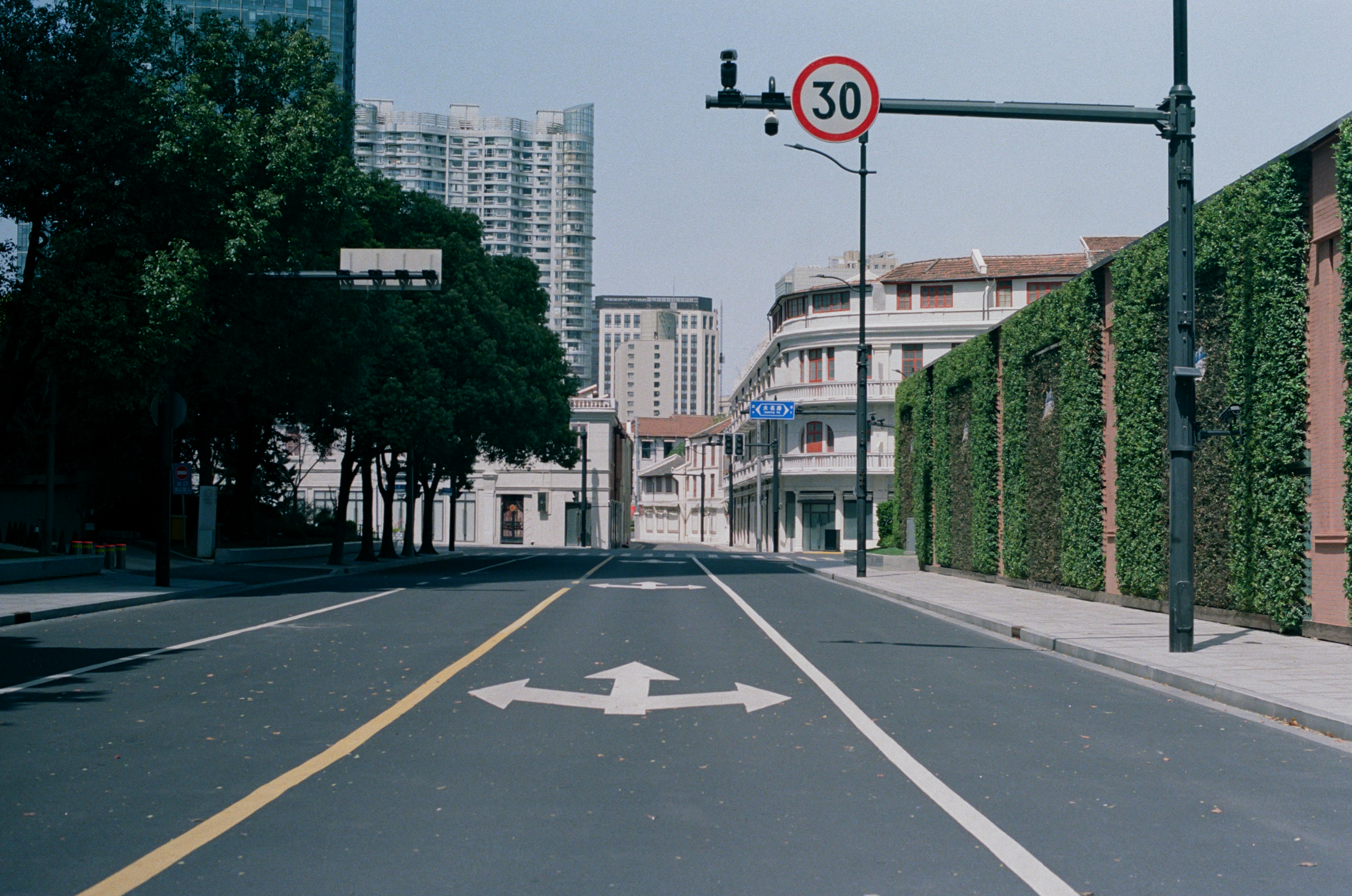 A quiet urban street featuring a roundabout, traffic signs, and buildings partially covered in greenery. The scene captures a blend of natural and architectural elements.