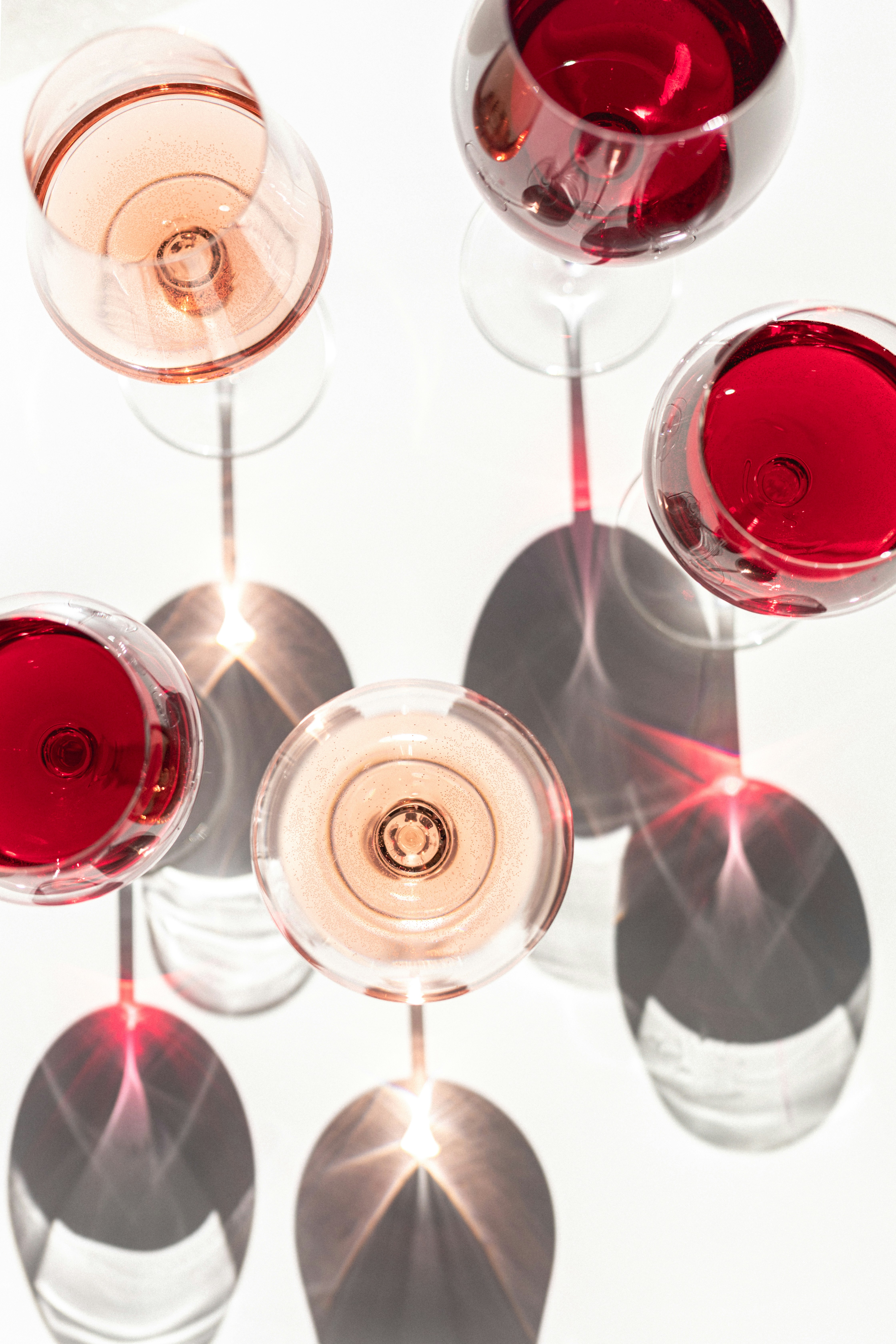 Five wine glasses arranged artistically, showcasing various shades of red and pink wines, casting intricate shadows on a white surface.