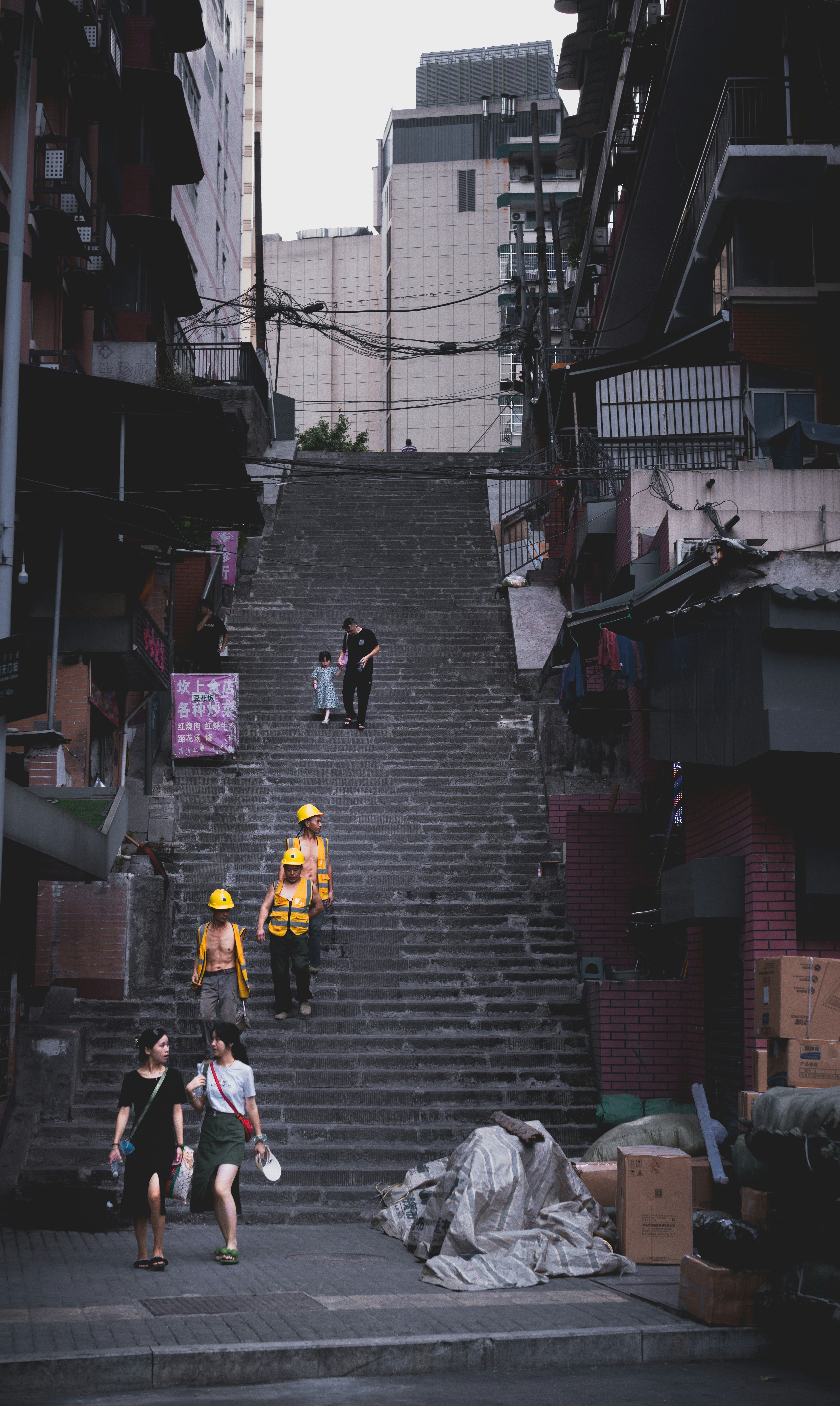 Concrete city stairway filled with pedestrians and a line of construction workers in yellow helmets descending between dense buildings and exposed wiring.