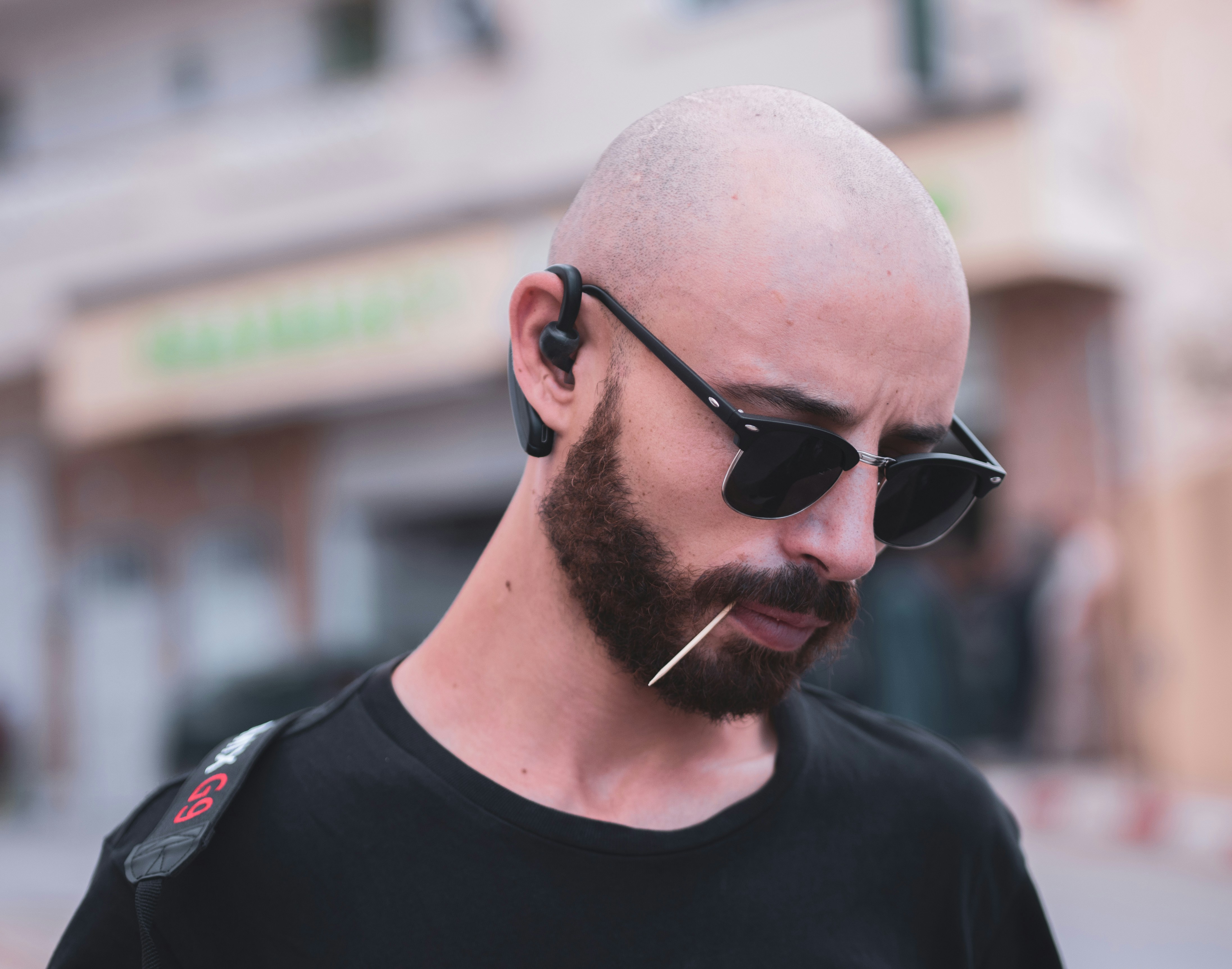 Bald man wearing sunglasses and earbuds, thoughtfully holding a toothpick in his mouth while standing in an urban setting.