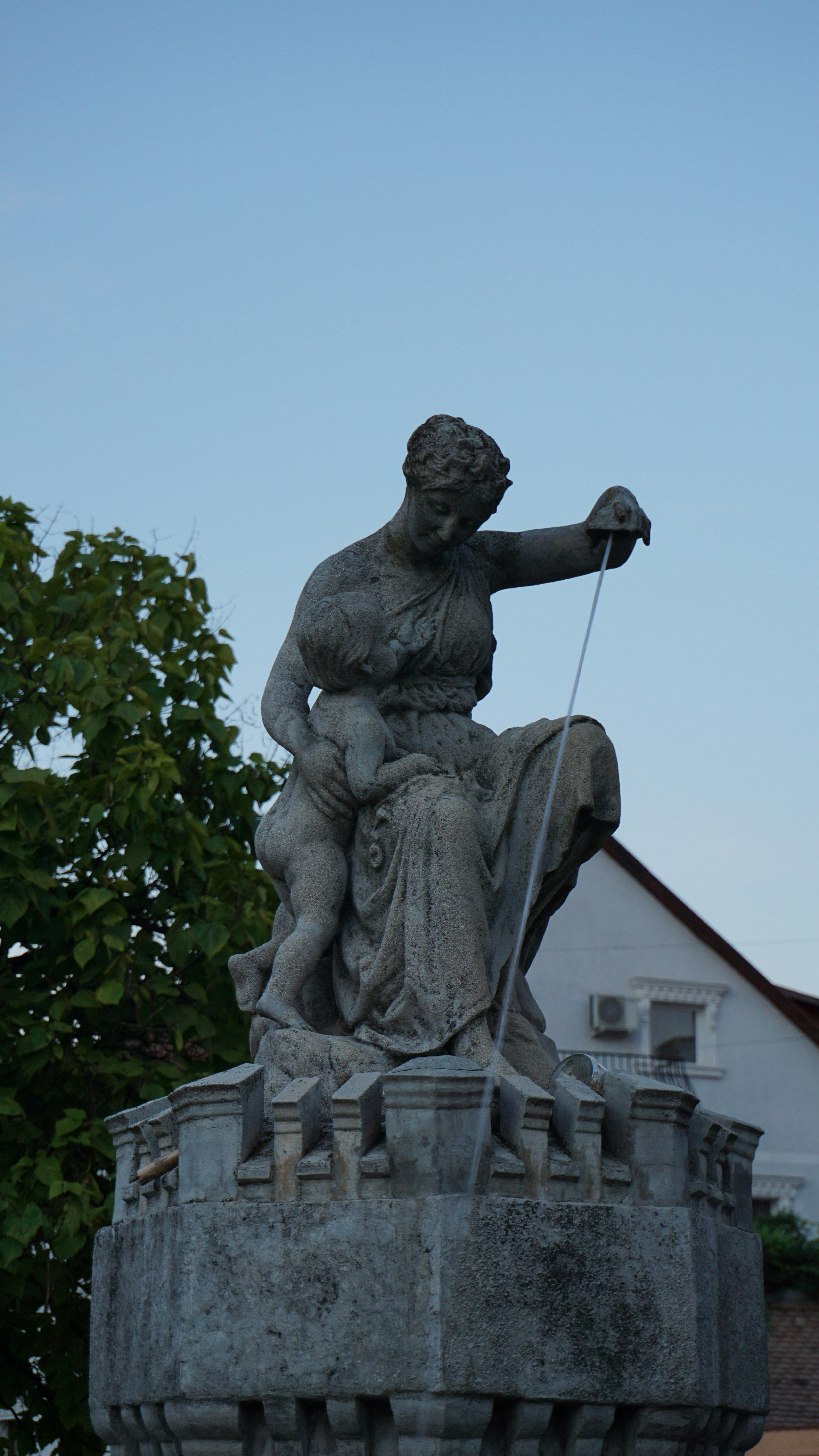A detailed statue of a woman holding a child, gracefully positioned atop a stone pedestal, surrounded by lush greenery and a clear sky.