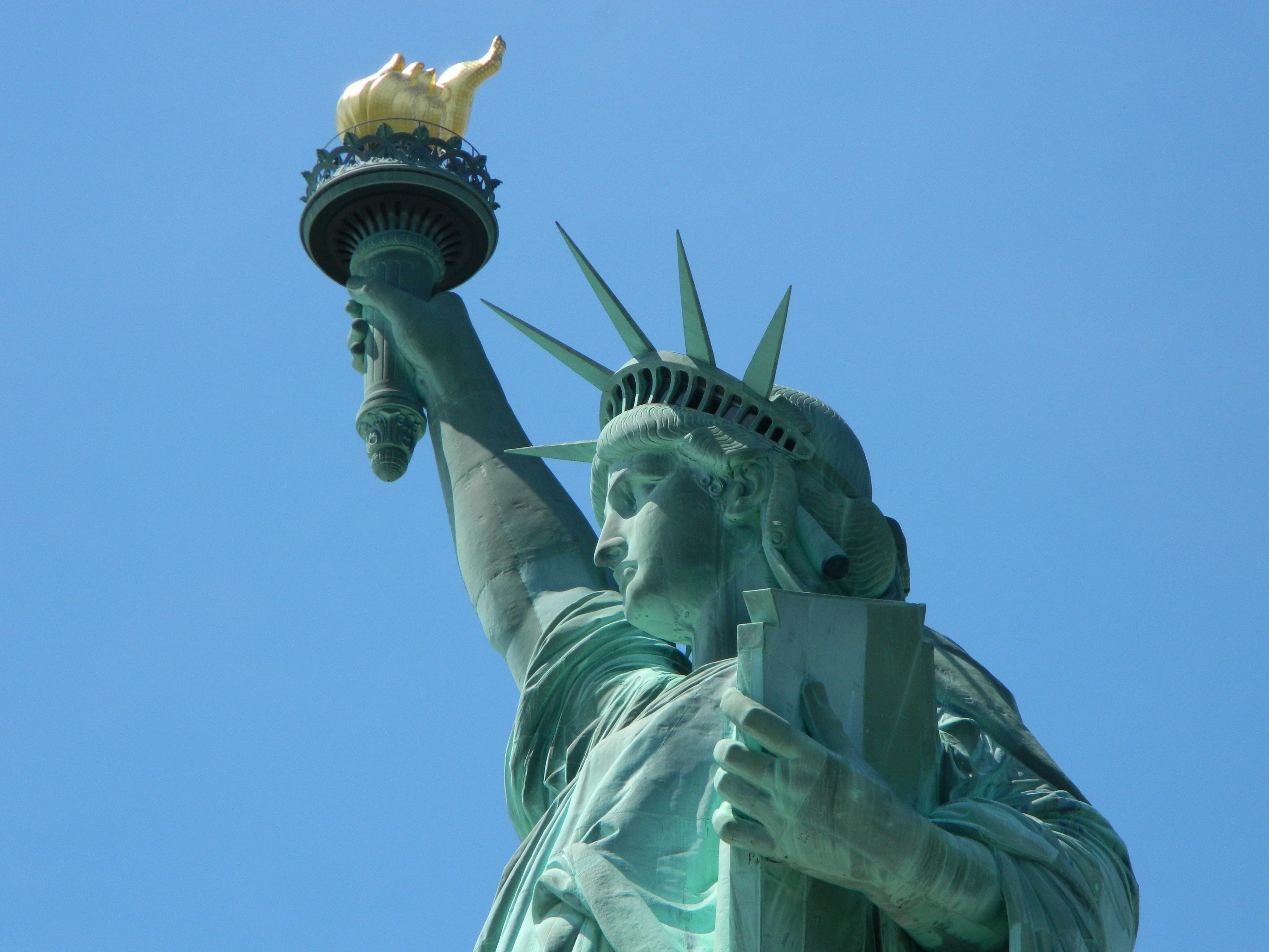 Statue of Liberty reaching upward with torch raised and tablet in close foreground against a bright blue sky. This is a photographic capture emphasizing iconic monument details.