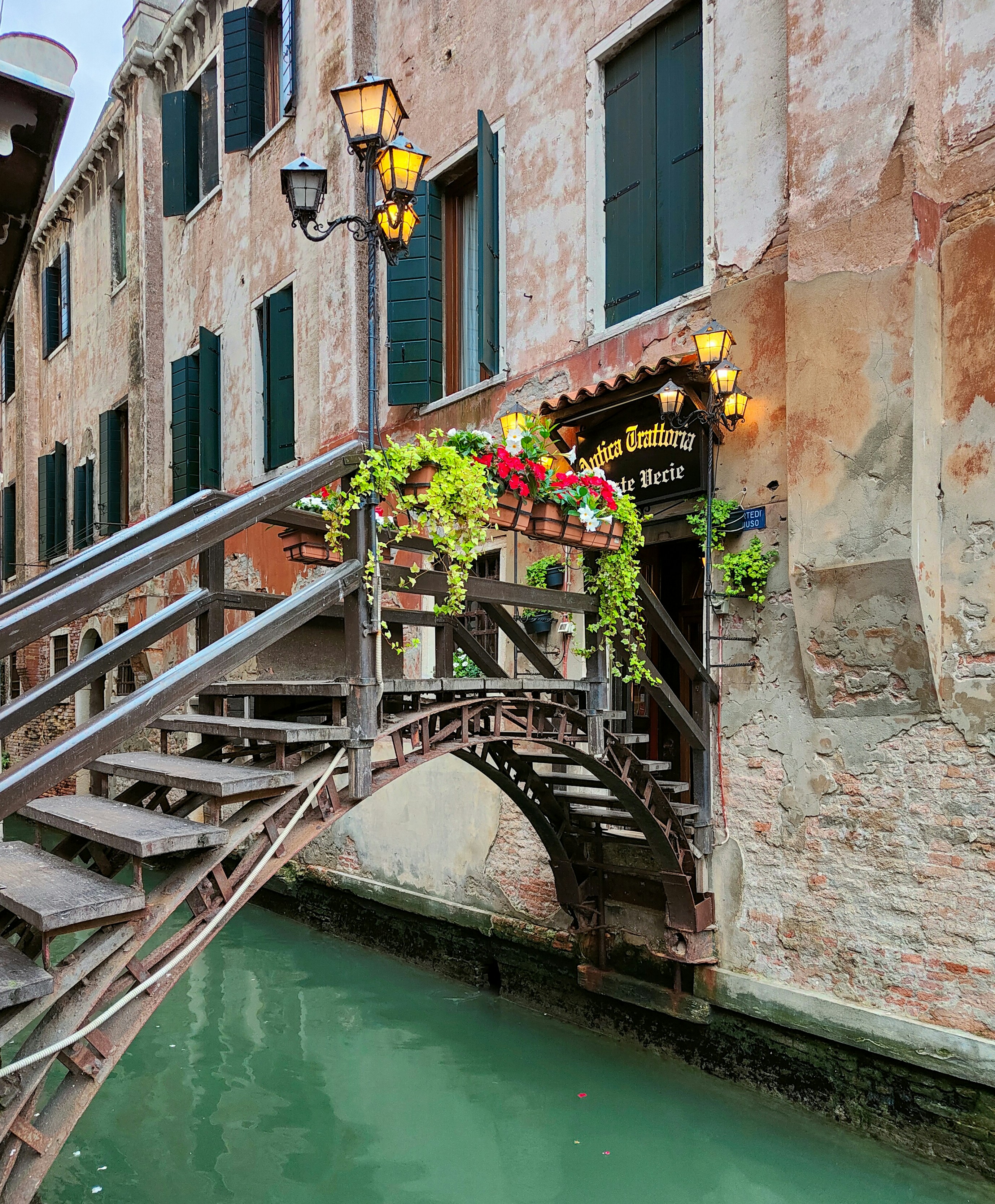 Brick canal-side building with a wooden arched bridge spanning a narrow green canal. Hanging planters and warm lanterns add color along the weathered facade.