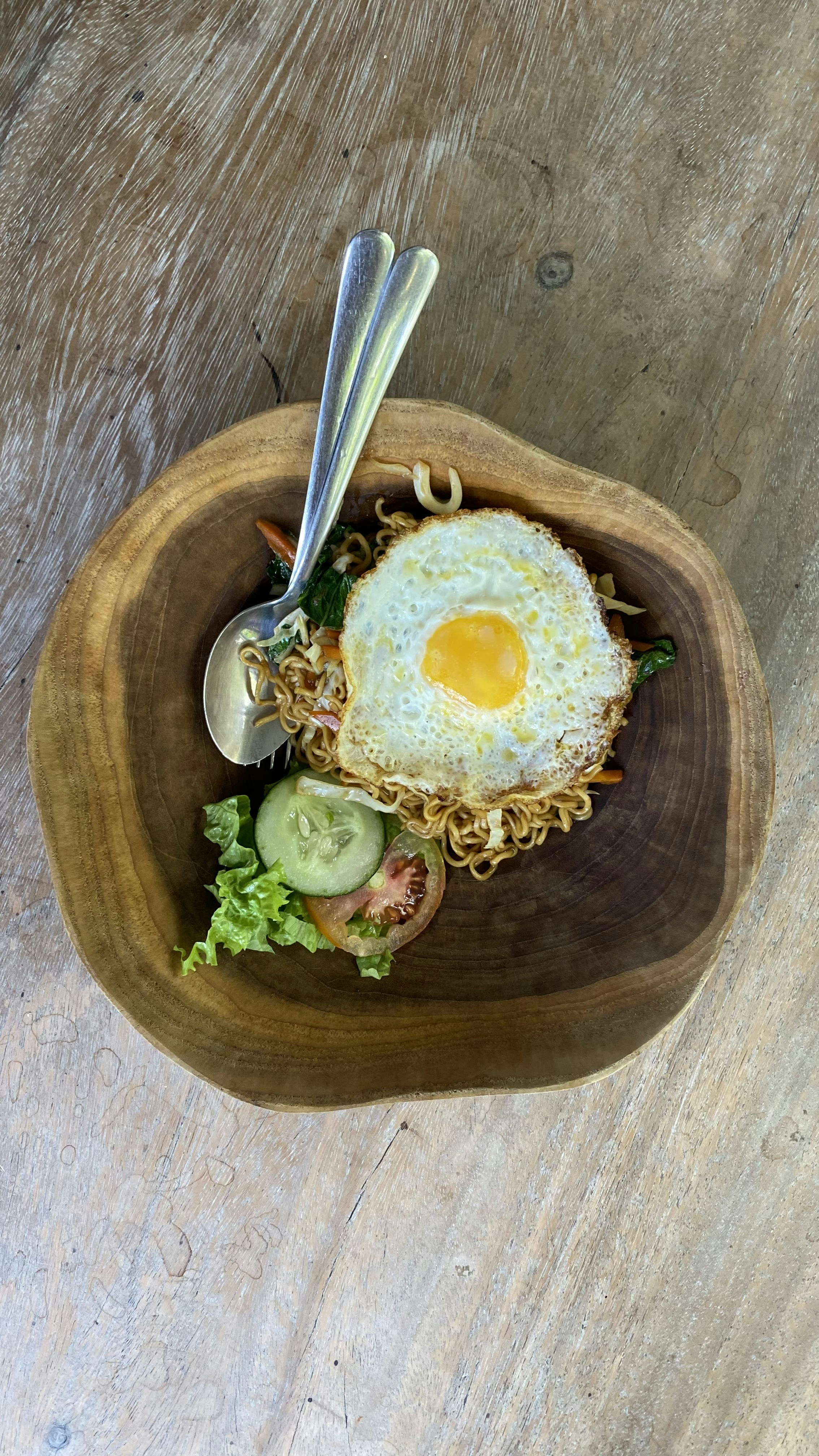 A wooden bowl filled with flavorful noodles topped with a sunny-side-up egg, accompanied by fresh vegetables and cutlery.