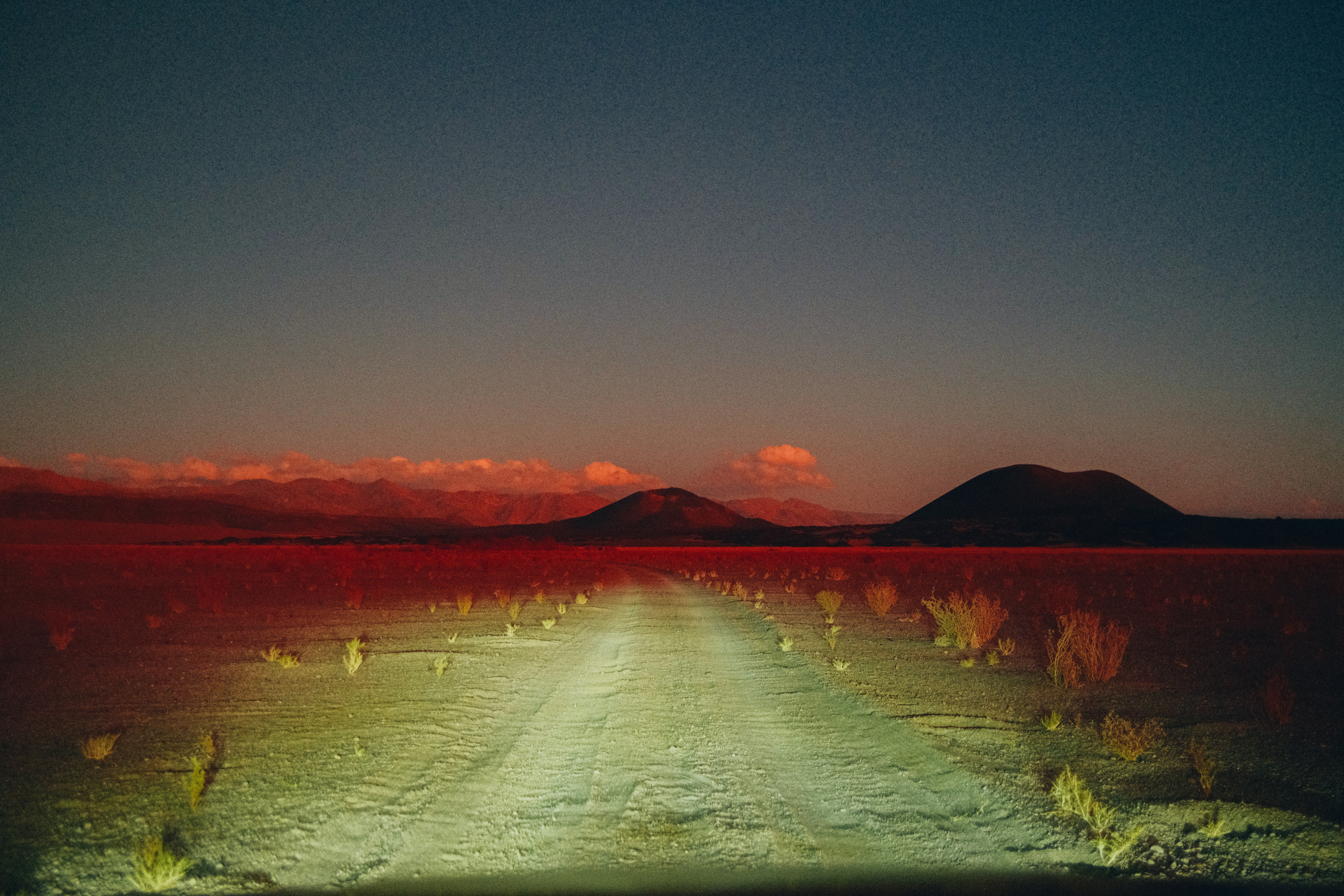 Dirt road leading to red desert horizon with volcano and mountain landscape with vegetation illuminated by vehicle headlights.