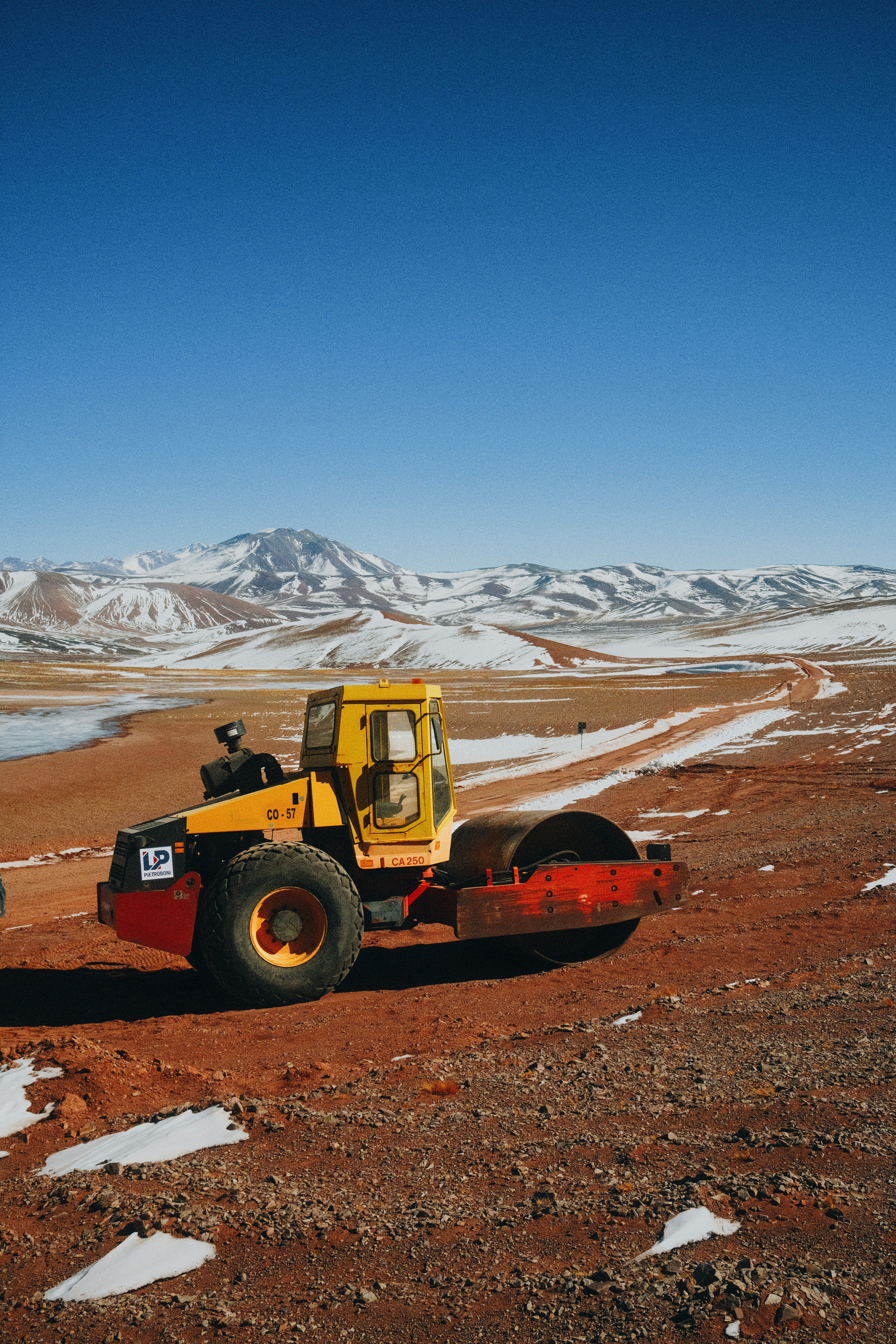 A yellow construction compactor sits on a snow-dusted red plain, with distant snow-covered mountains under a blue sky.