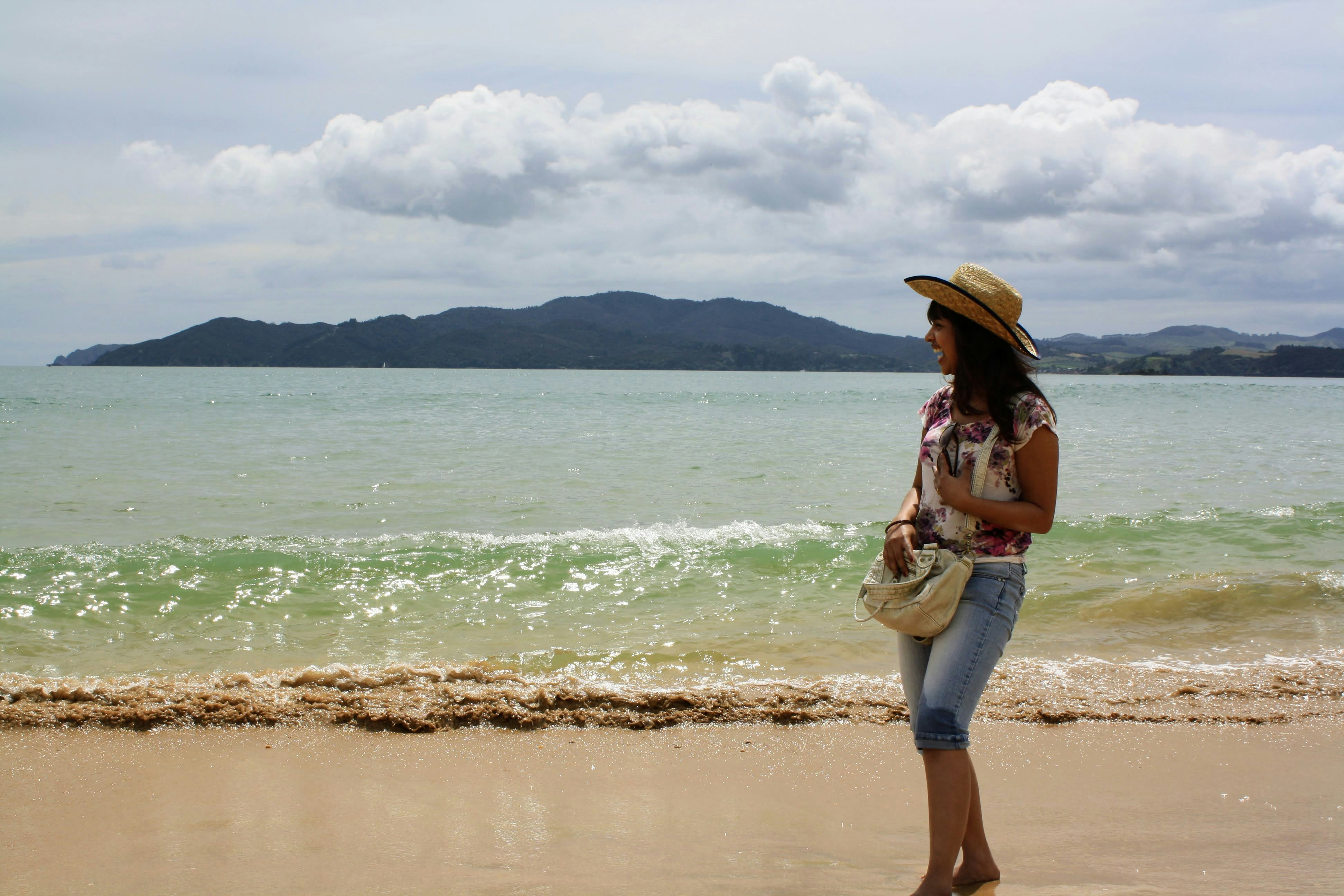Woman in a sun hat standing on a beach, gazing towards distant hills under a cloudy sky.