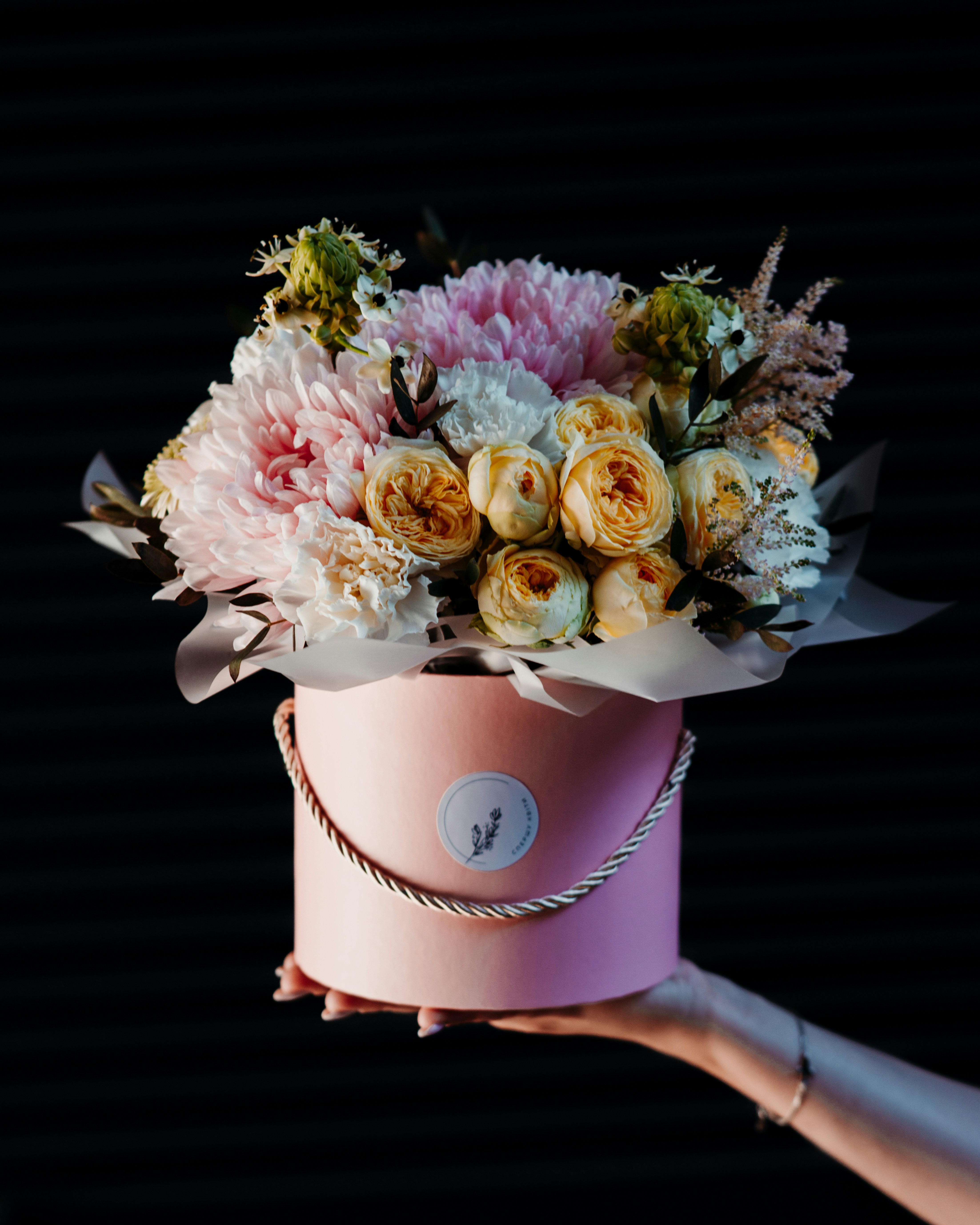 A delicate arrangement of pastel roses and chrysanthemums in a pink box, held gracefully against a dark backdrop.