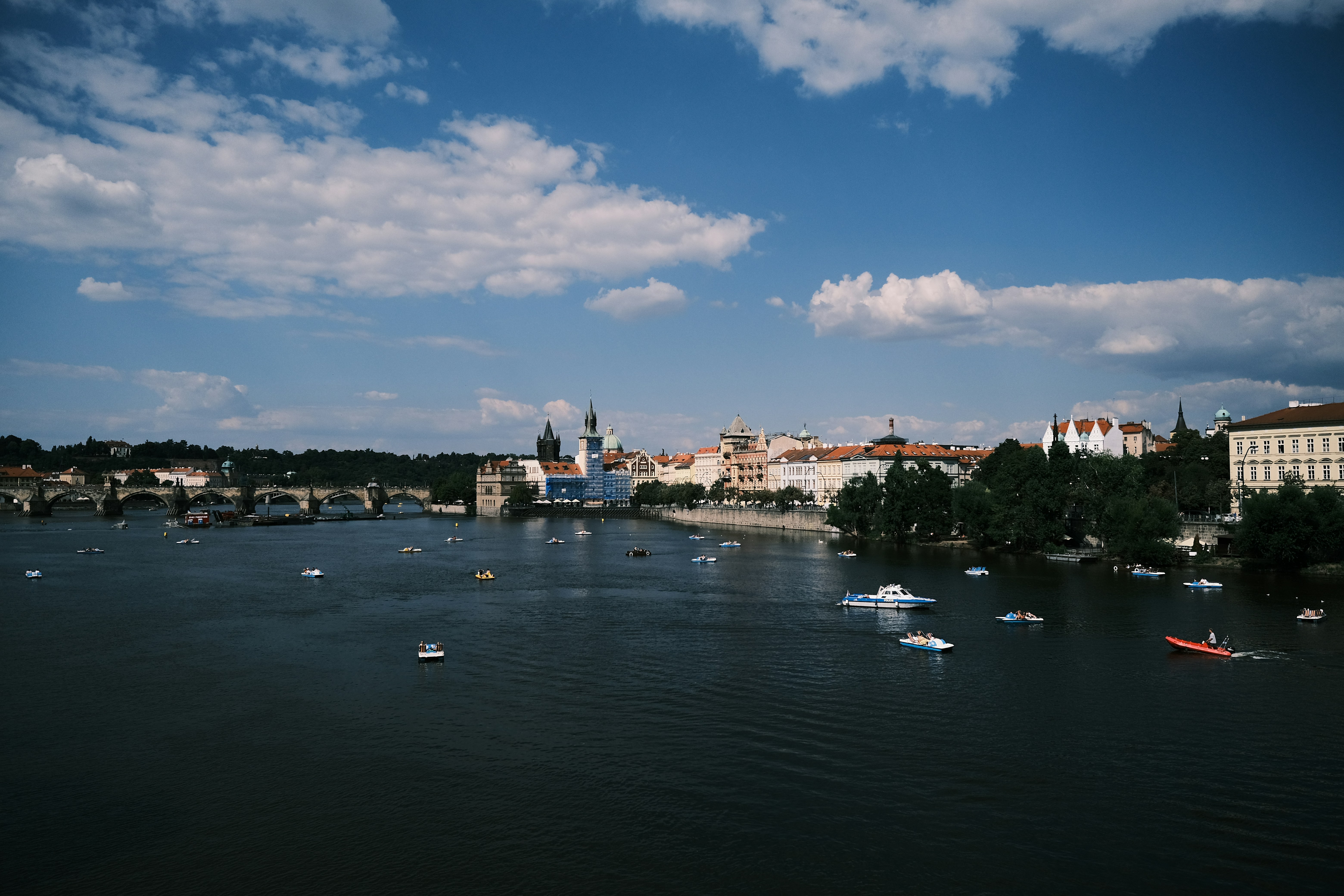 Vibrant boats navigate the tranquil waters of the Vltava River, framed by the historic architecture of Prague under a bright blue sky.