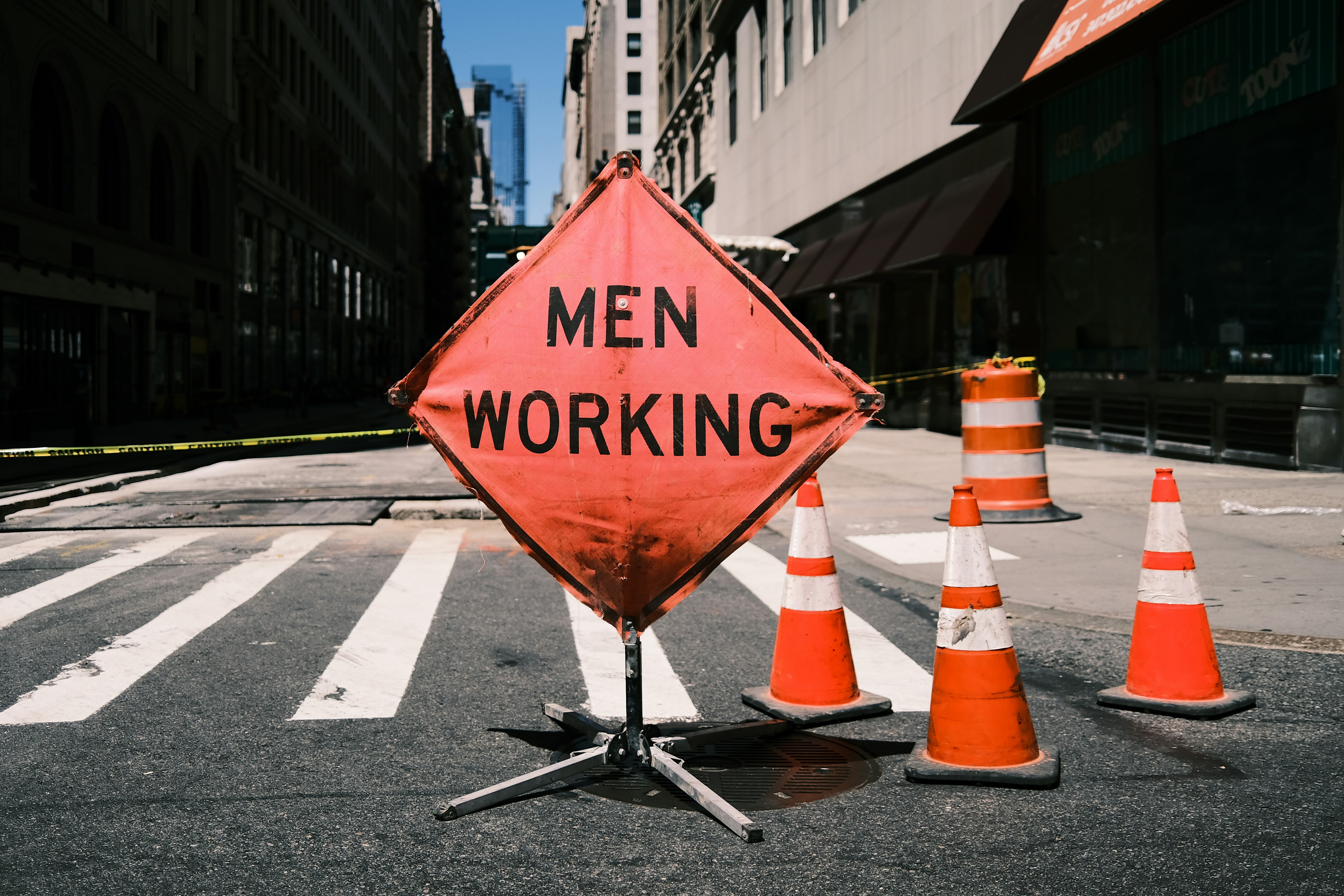 Bright orange construction sign reading 'MEN WORKING' on city street, surrounded by traffic cones. The scene captures the essence of urban development.