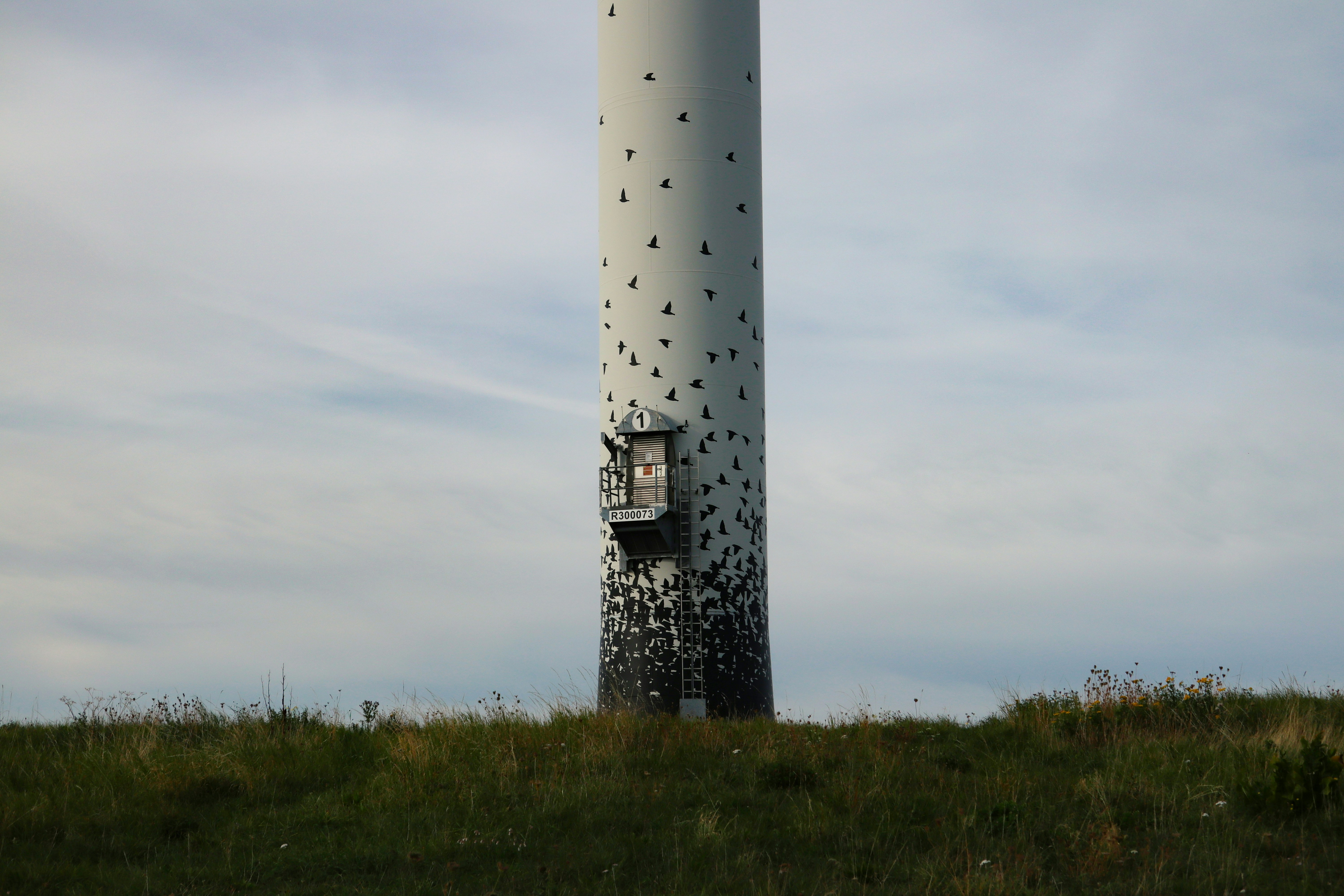 A wind turbine tower adorned with black silhouettes, contrasting sharply against a cloudy sky, showcases a unique artistic interpretation of nature and technology.