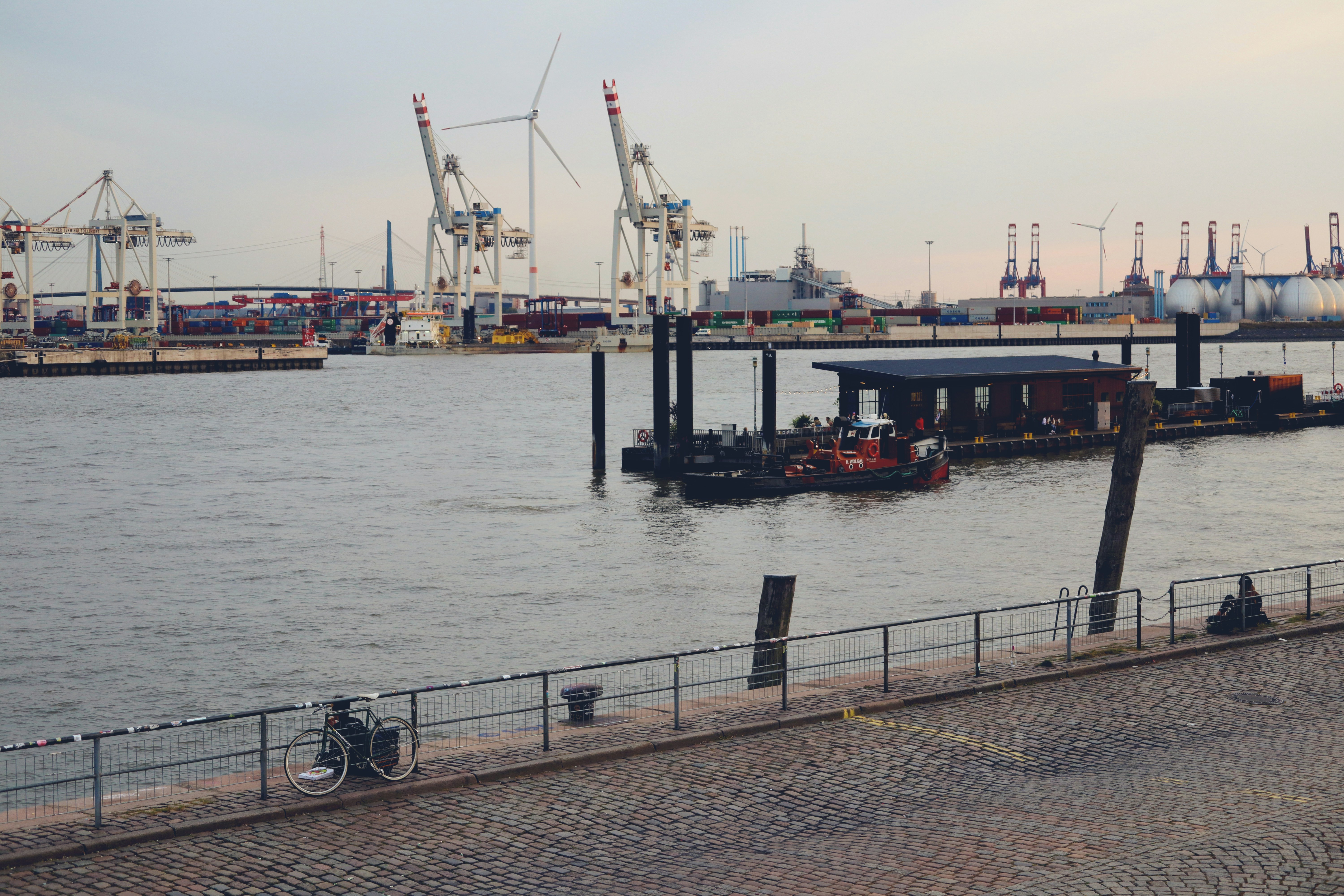 Industrial harbor scene featuring boats and cranes with a calm water surface reflecting the sky.
