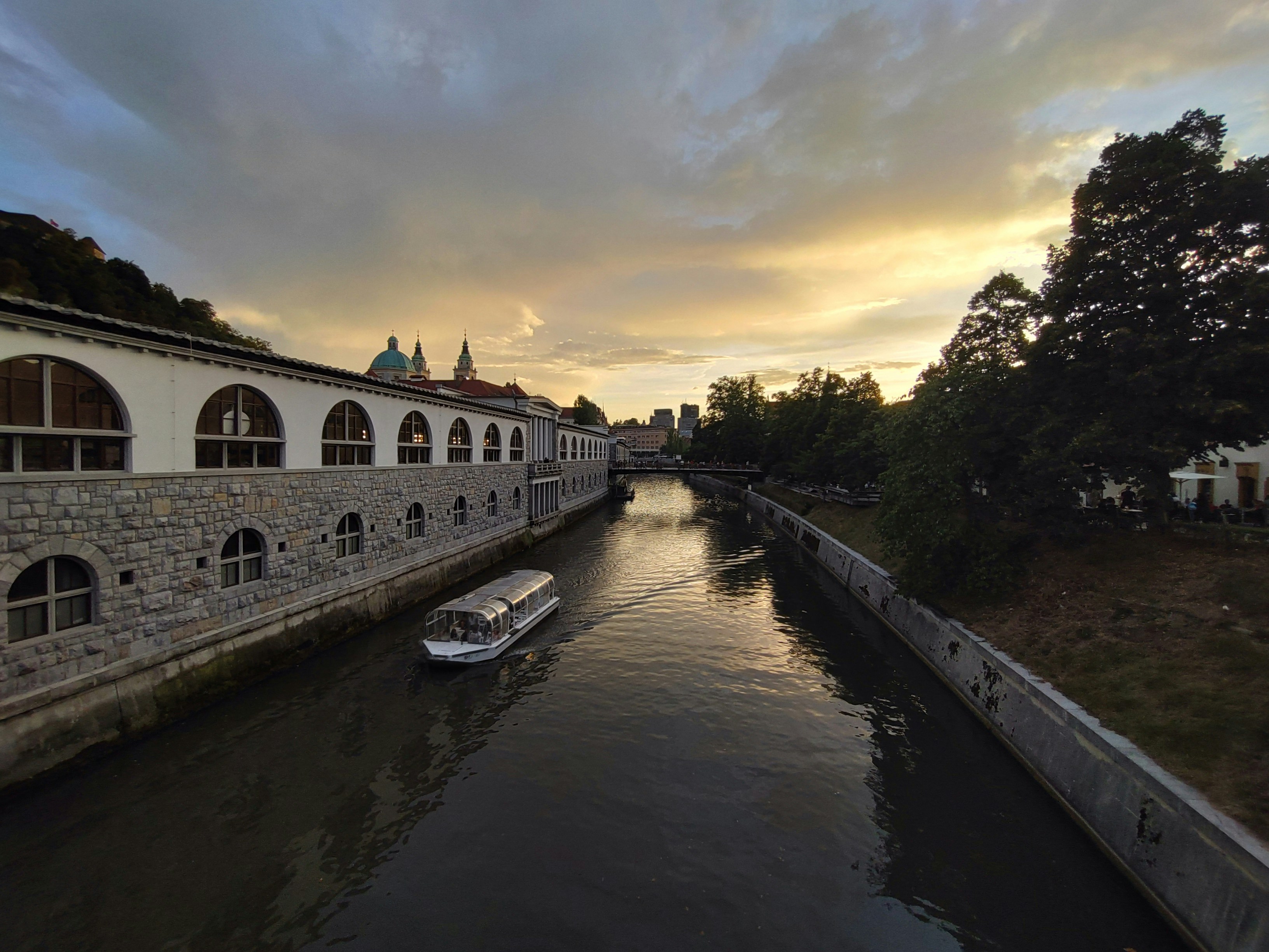 Sunset light bathes a canal flanked by a long stone building with arched windows as a small boat glides along the water. Trees line the opposite bank while clouds glow above.