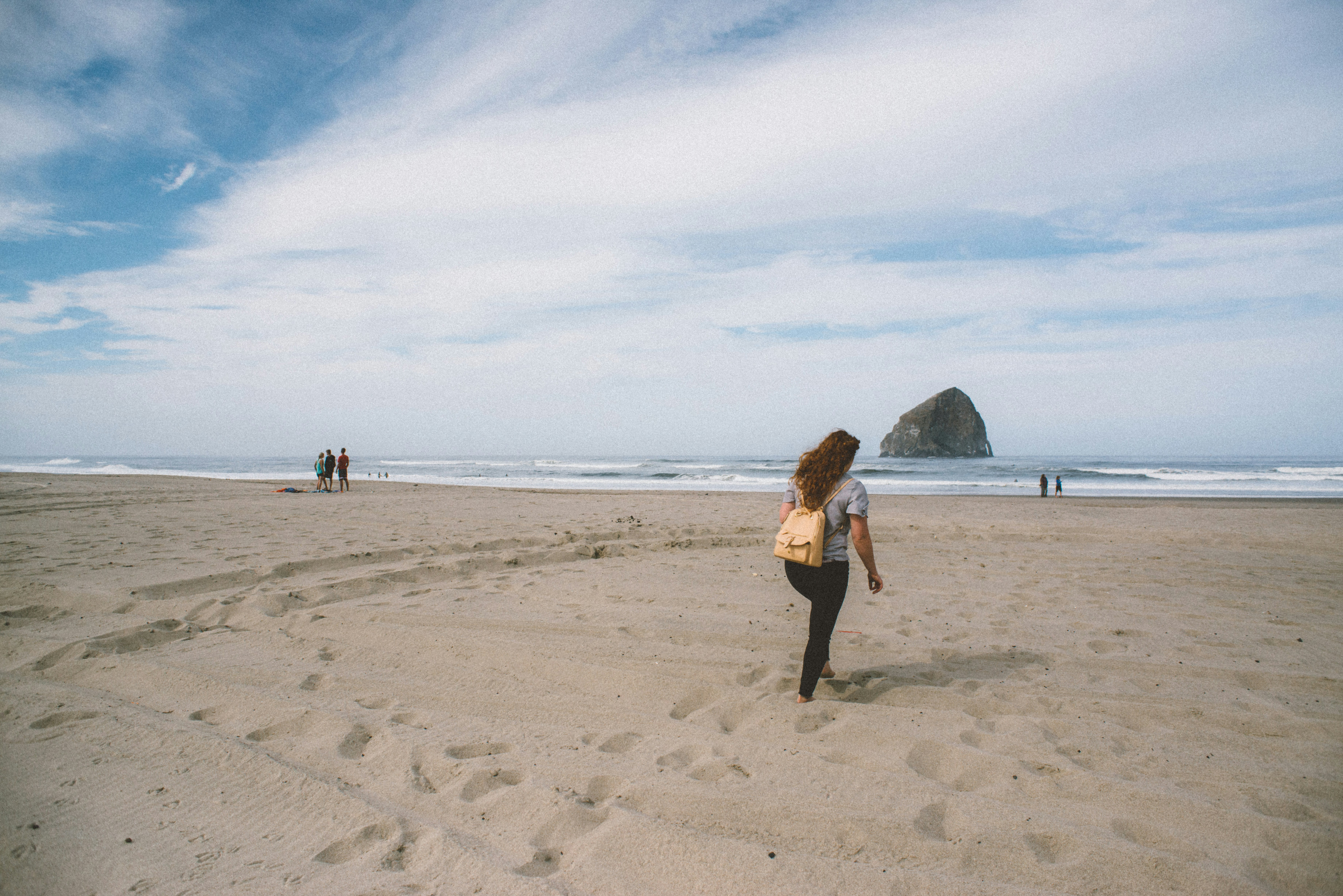 Girl with backpack on a windswept beach approaching a distant rock