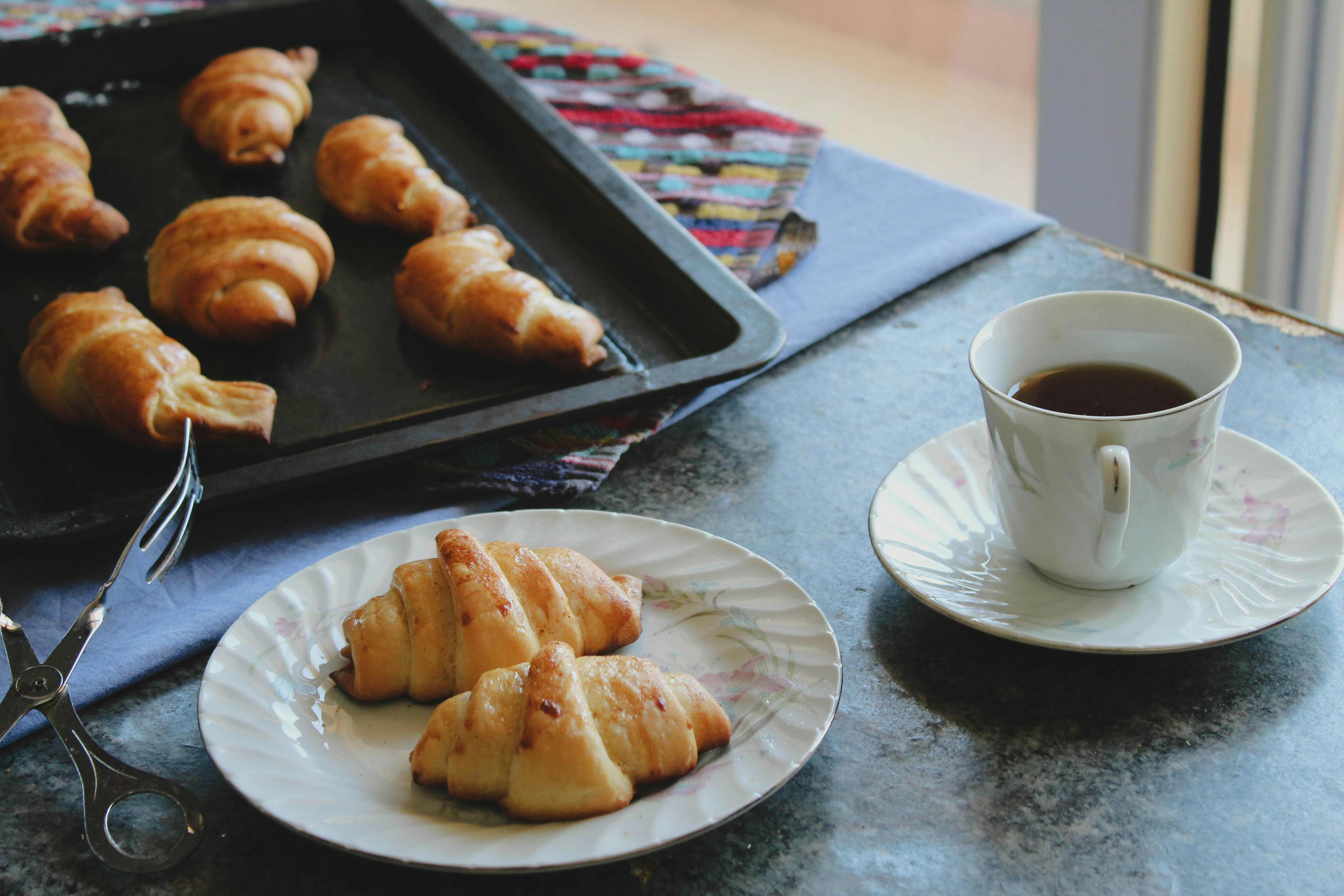 Golden croissants on a plate and baking tray beside a steaming cup of coffee on a rustic table.